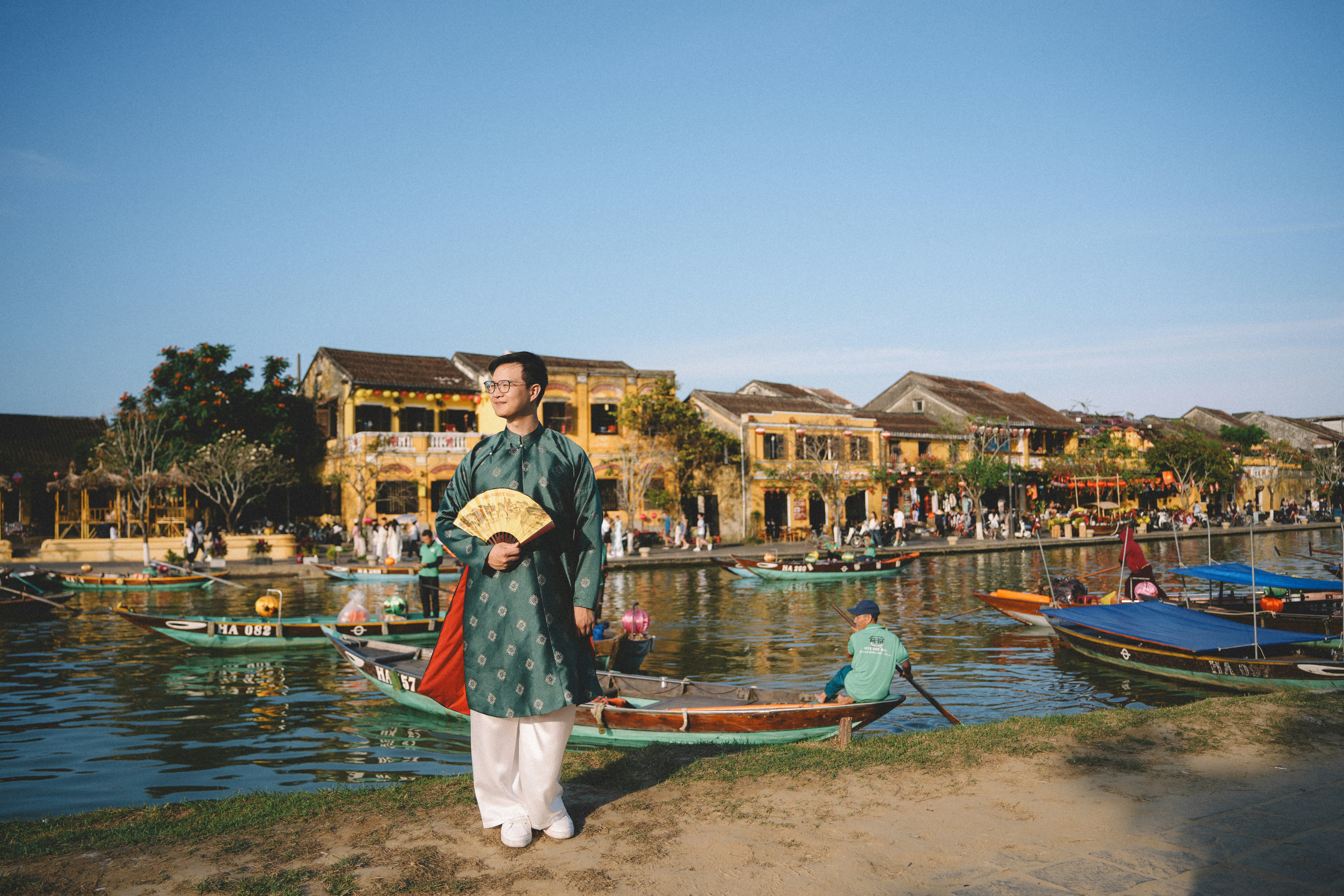 Portrait in a riverside bazaar: man in green robe with fan as boats drift behind