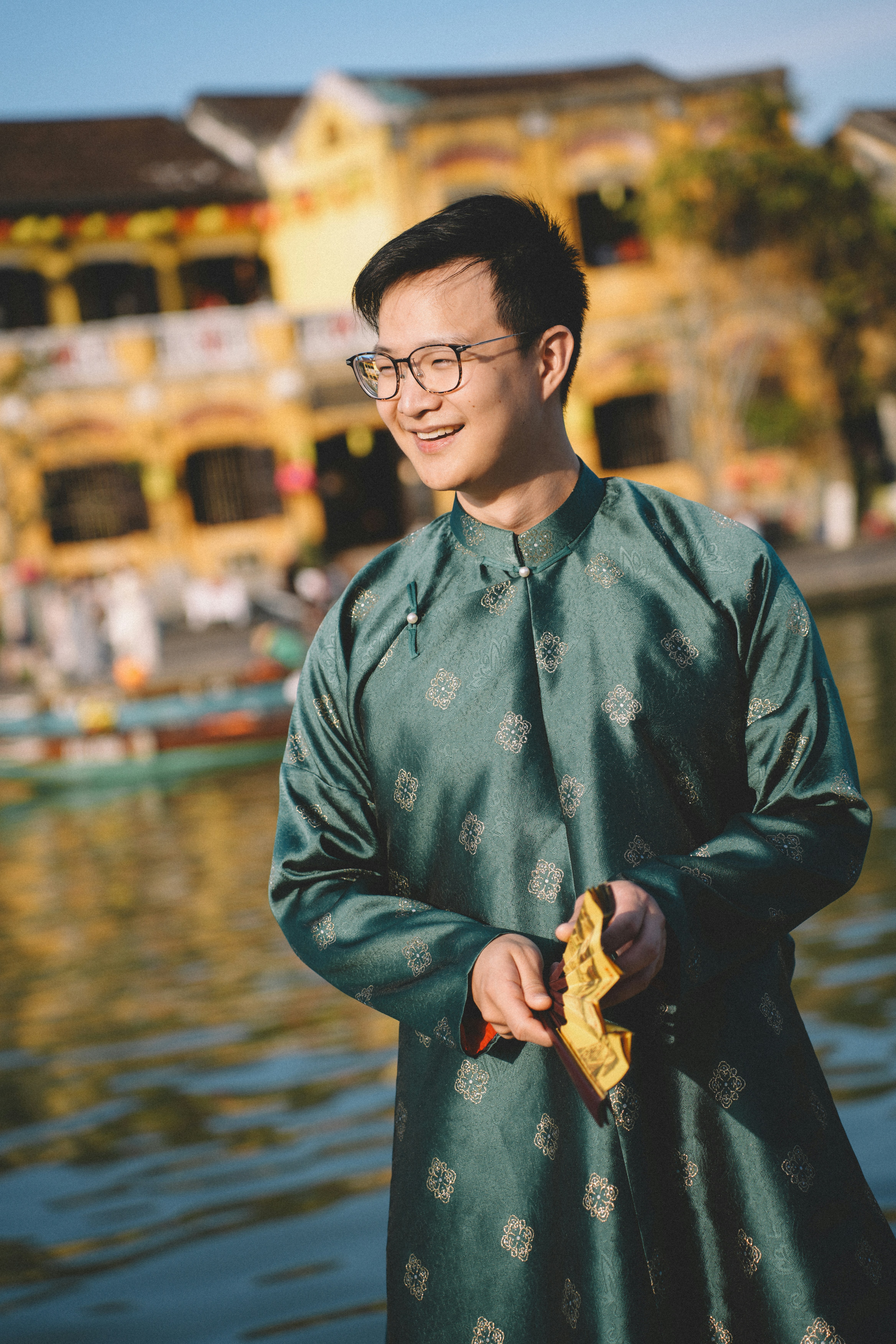 Gentleman in teal áo dài smiles beside sunlit canal at a colorful riverside