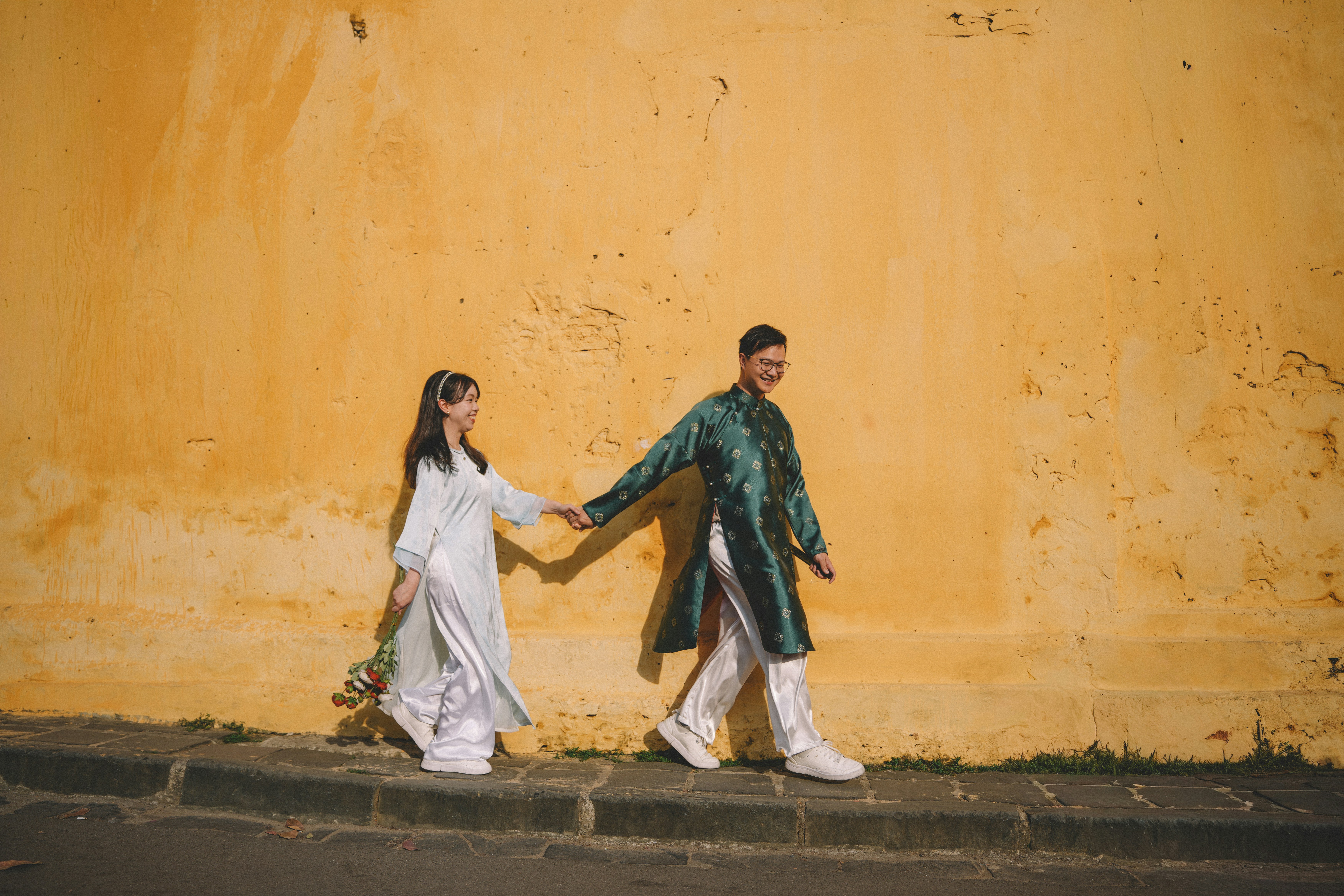 Couple in Traditional Attire Walks Along a Sunlit Yellow Wall