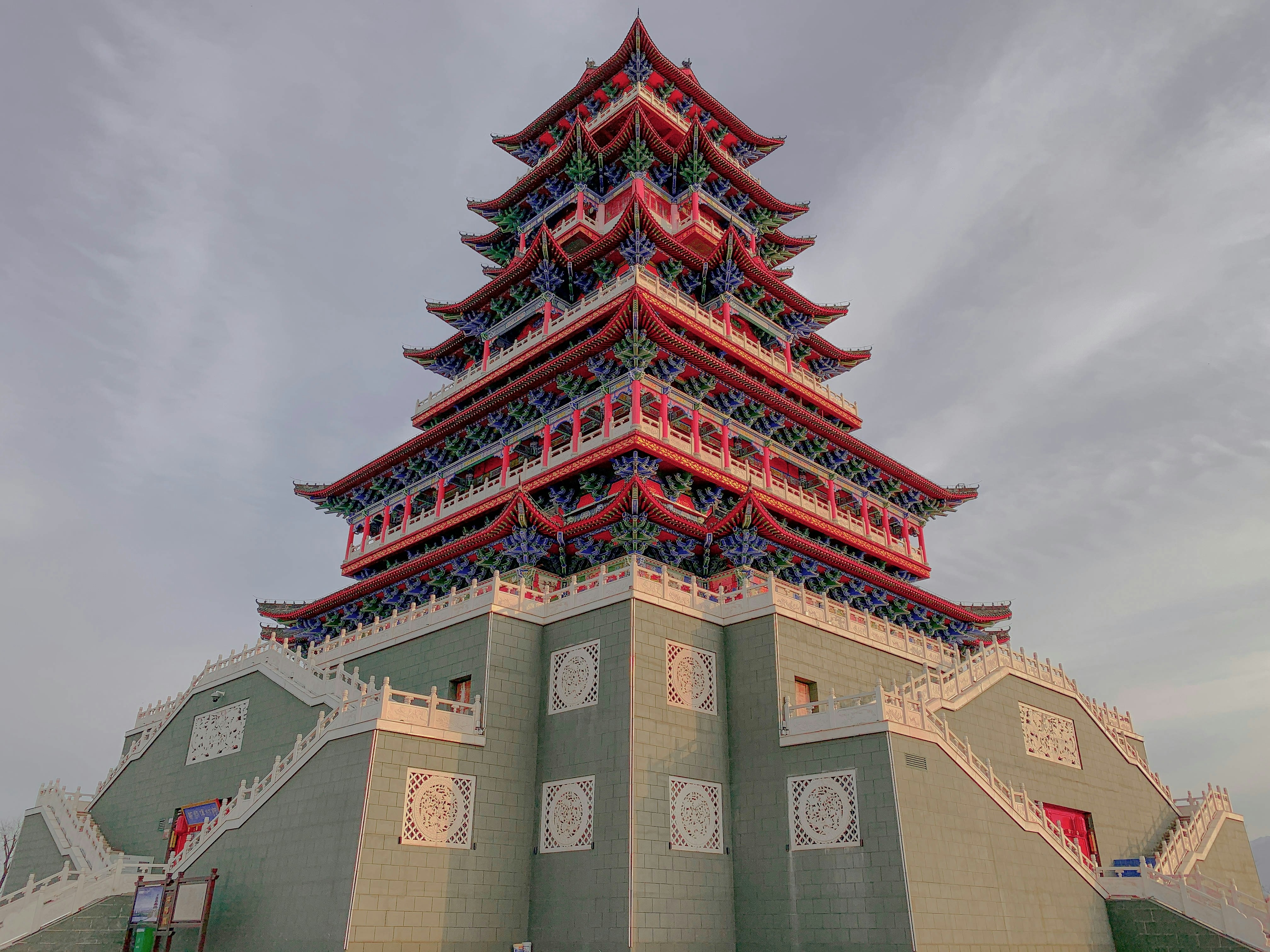 Tall, ornate pagoda perched on a broad, stair-stepped base with pastel-green walls and decorative lattice panels against a soft sky.