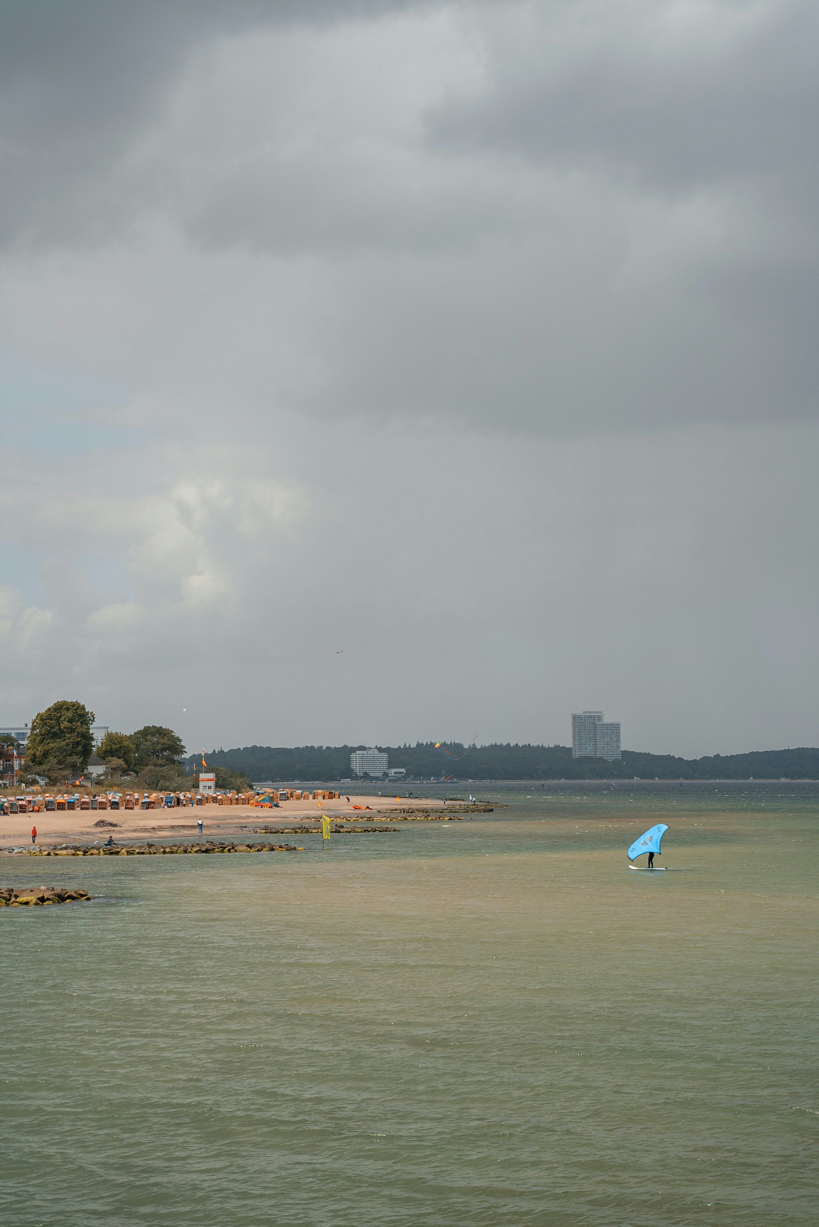 Muted coastal scene with a distant windsurfer and a calm shoreline under heavy clouds. A hint of human activity along the beach provides context for scale and mood.