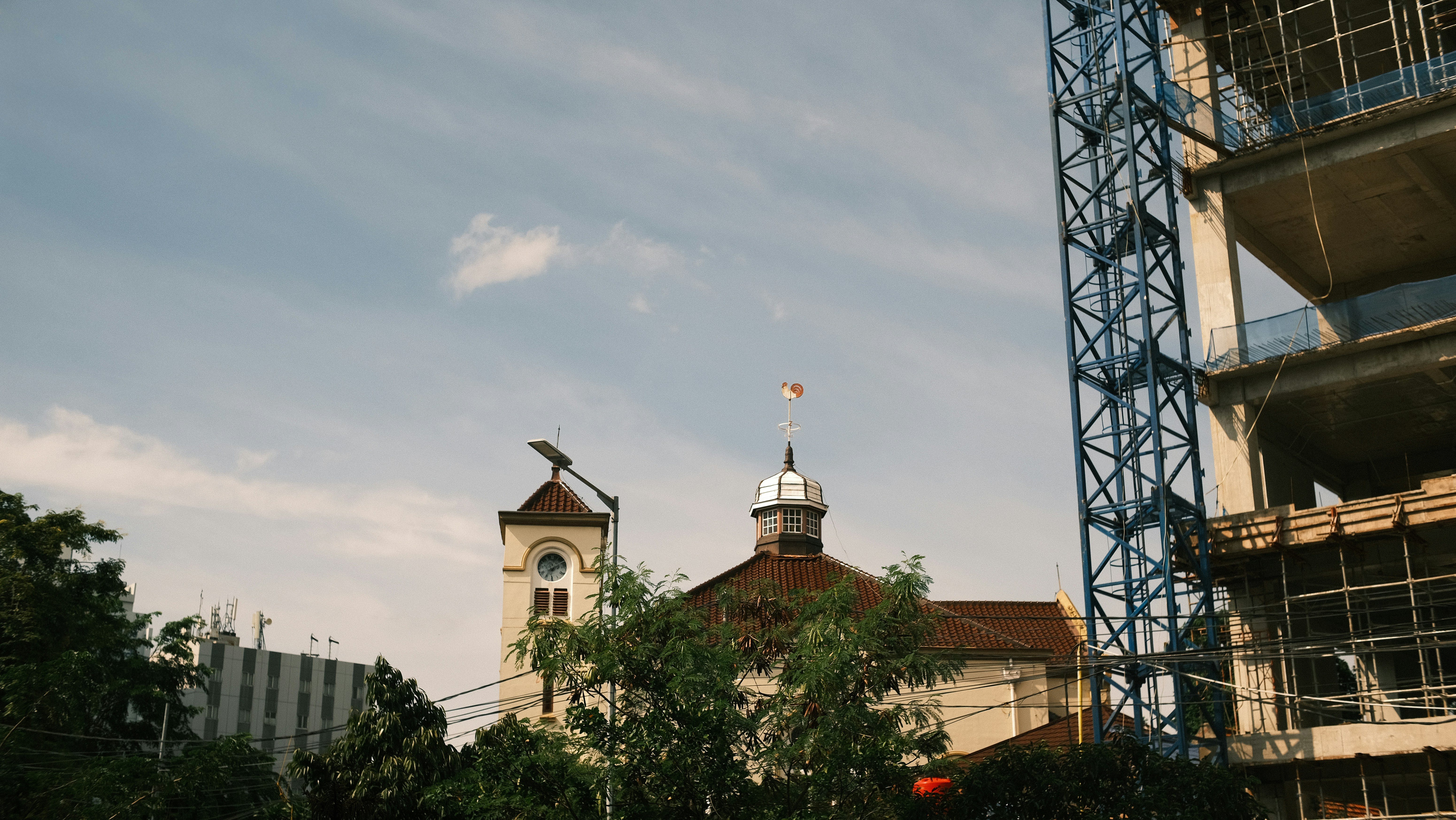 A street-level view of a historic building with a clock tower partially obscured by trees, framed by a construction scaffold on the right; the scene captures a moment of architectural contrast in daylight. The image appears to be a photograph rather than a painting or illustration.