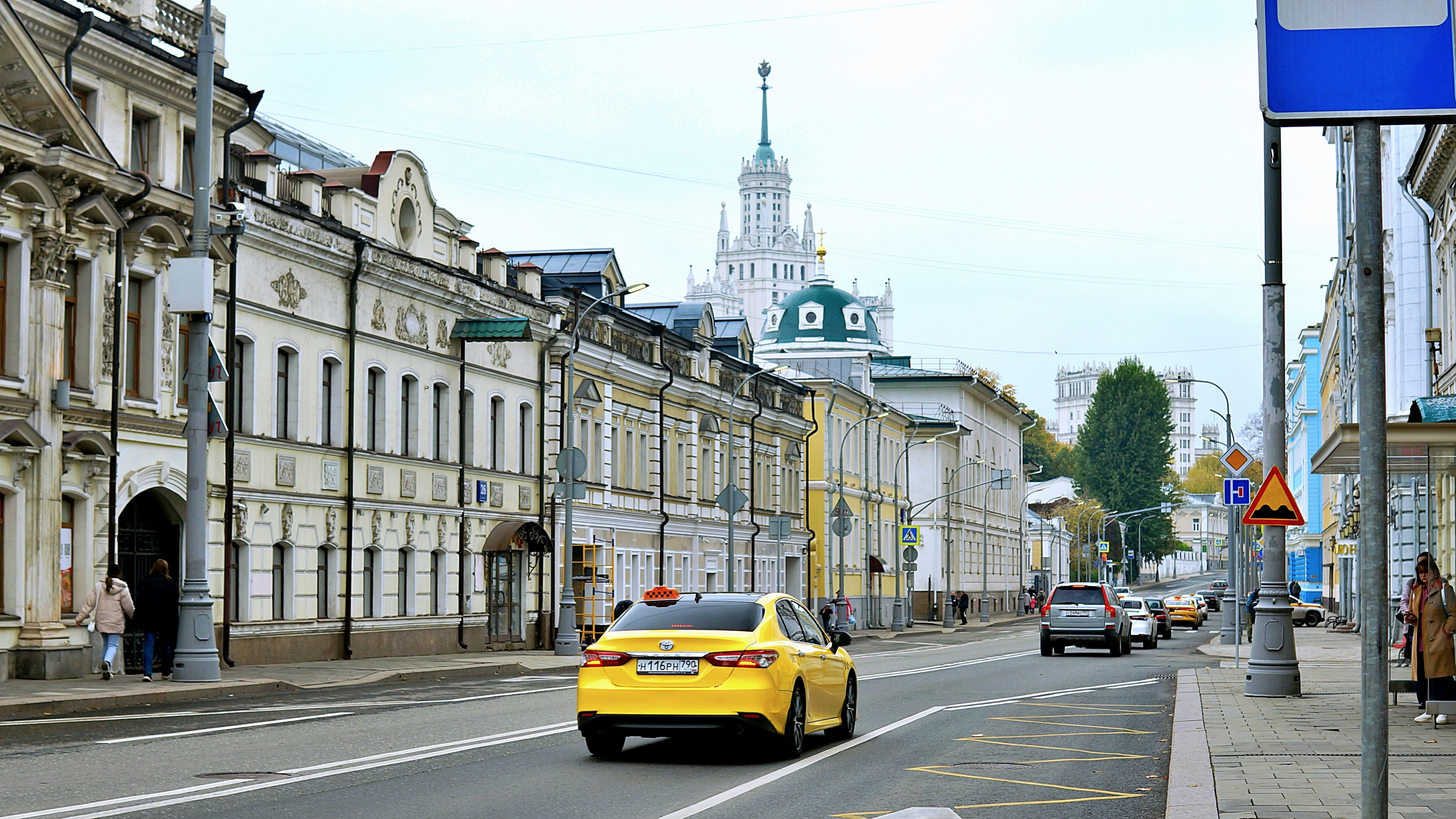 A yellow taxi drives down a sunlit historic street lined with pale, ornate buildings and a distant tower, capturing urban motion and architectural detail. This is a street photography scene with real-world urban context.