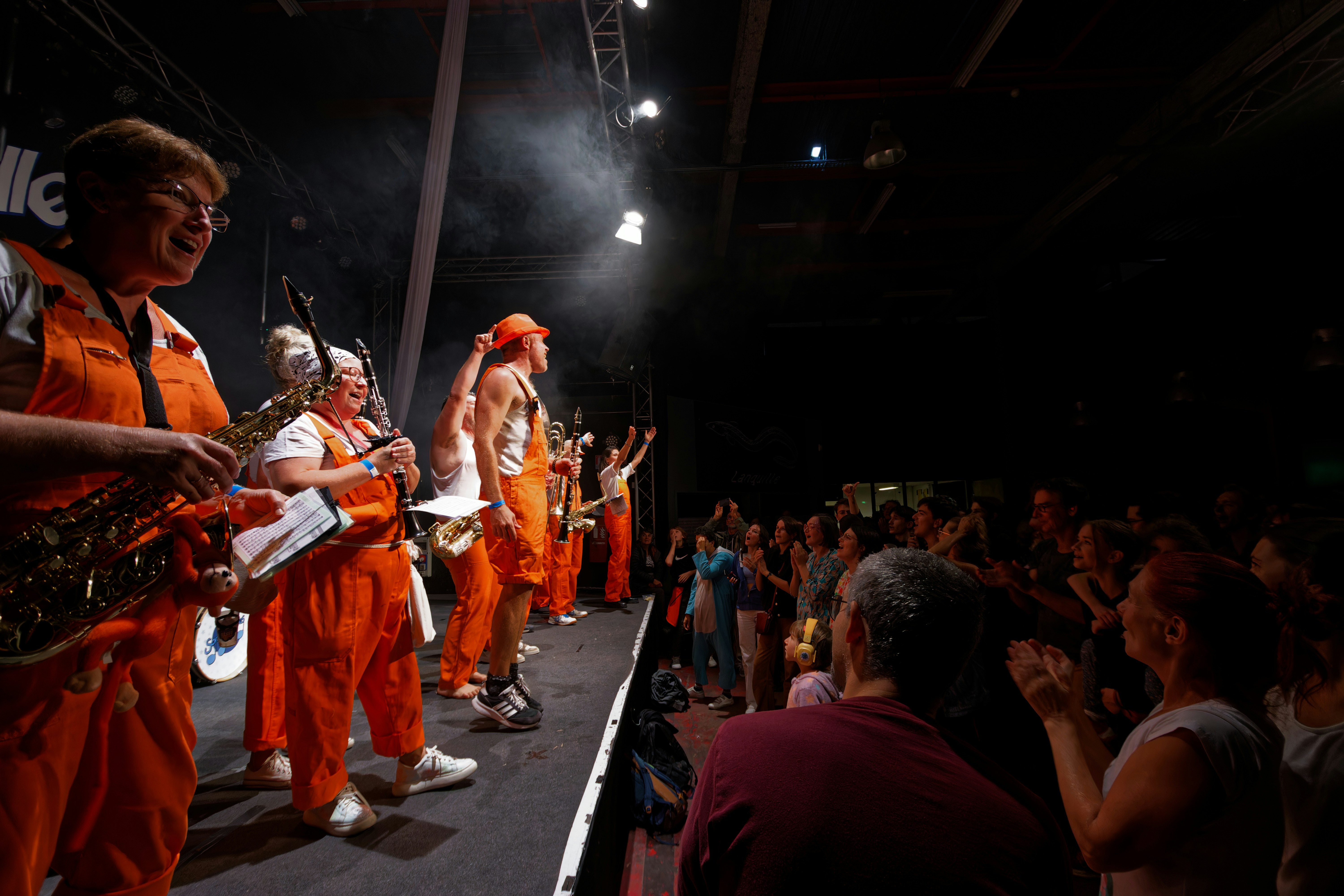 Orange-clad band on stage saluting a cheering crowd under stage lights