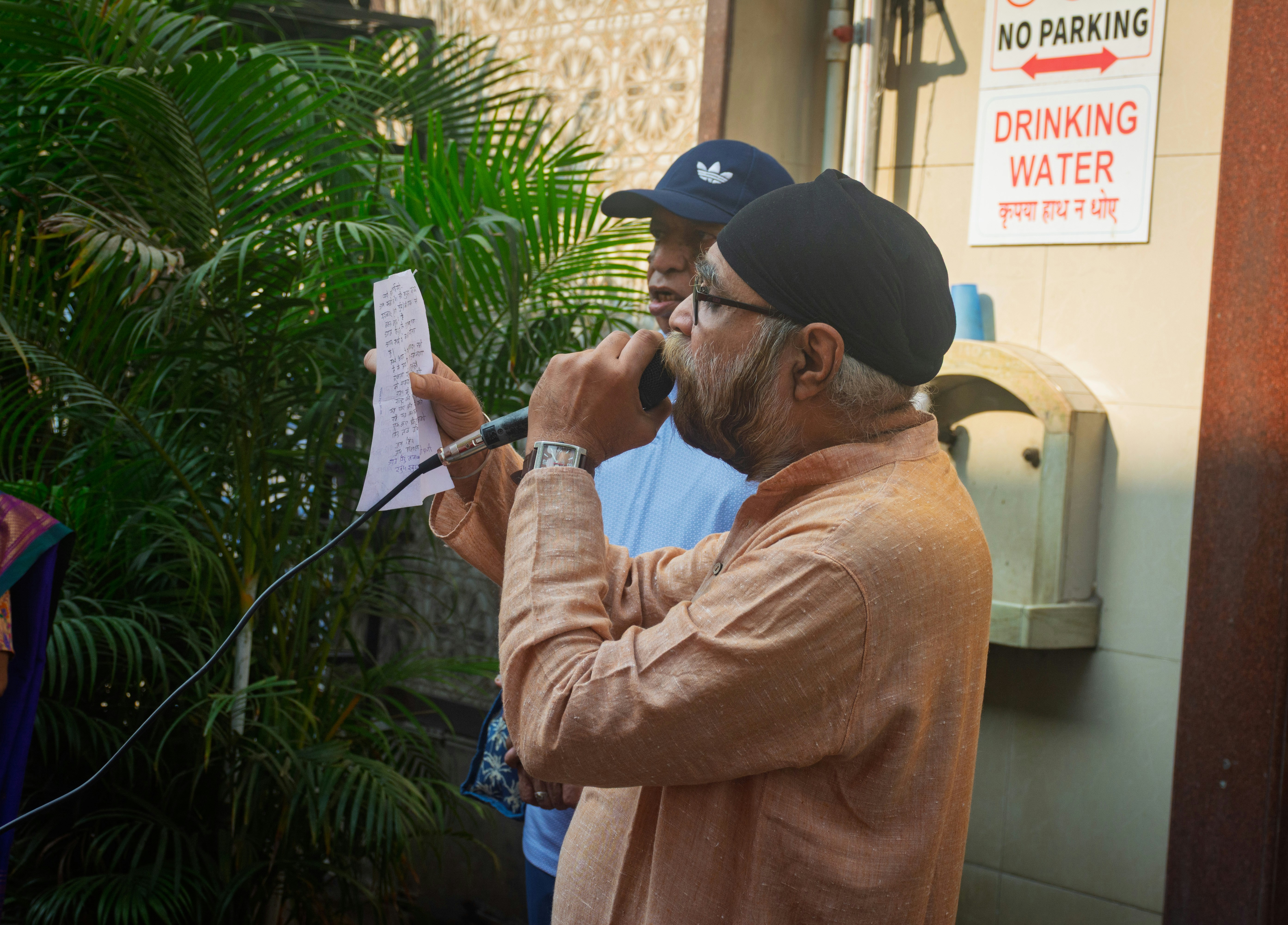 Speaker in a saffron shirt delivers notes amid greenery and a wall of signage