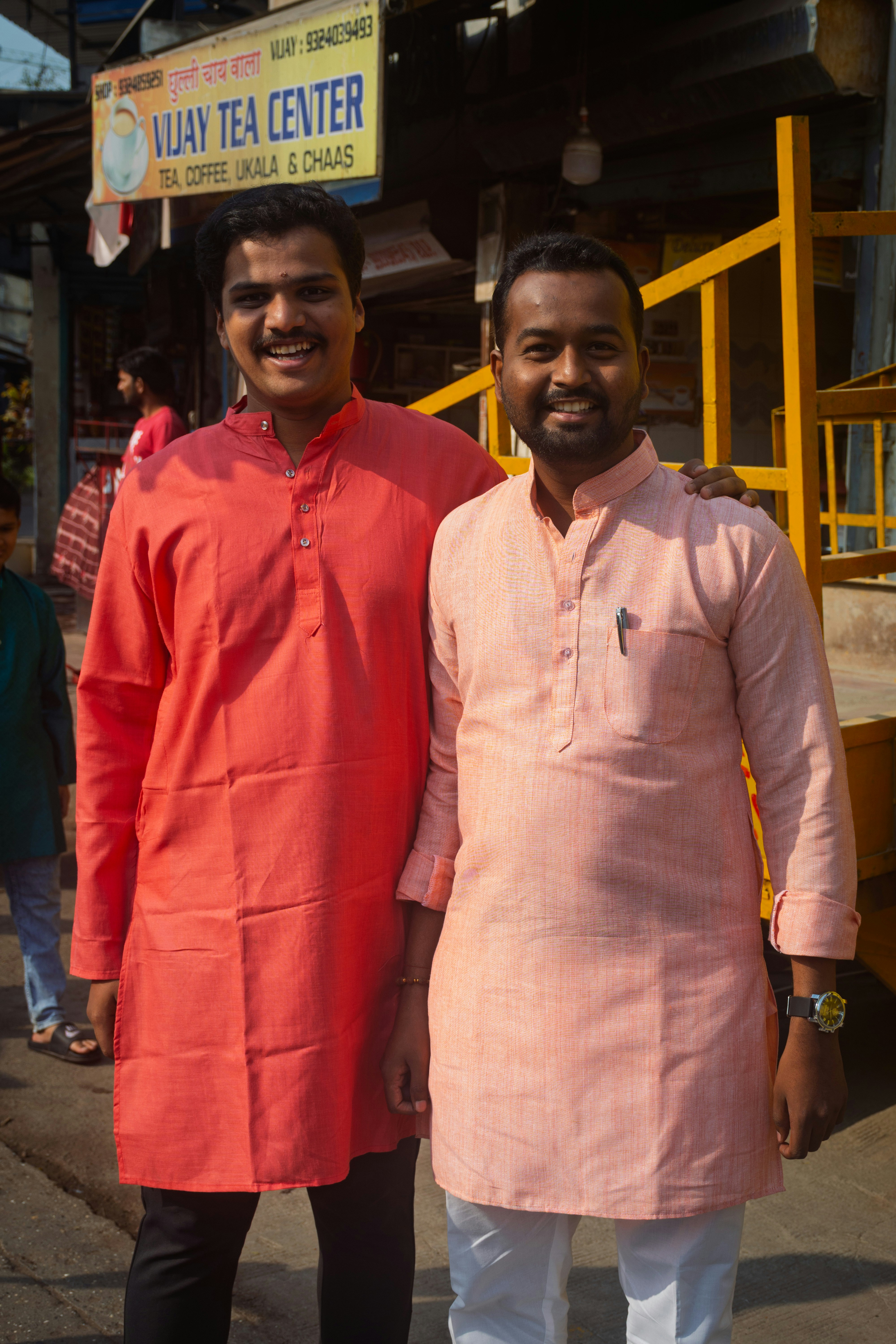 Two friends in vibrant kurta stand smiling beside a tea shop sign