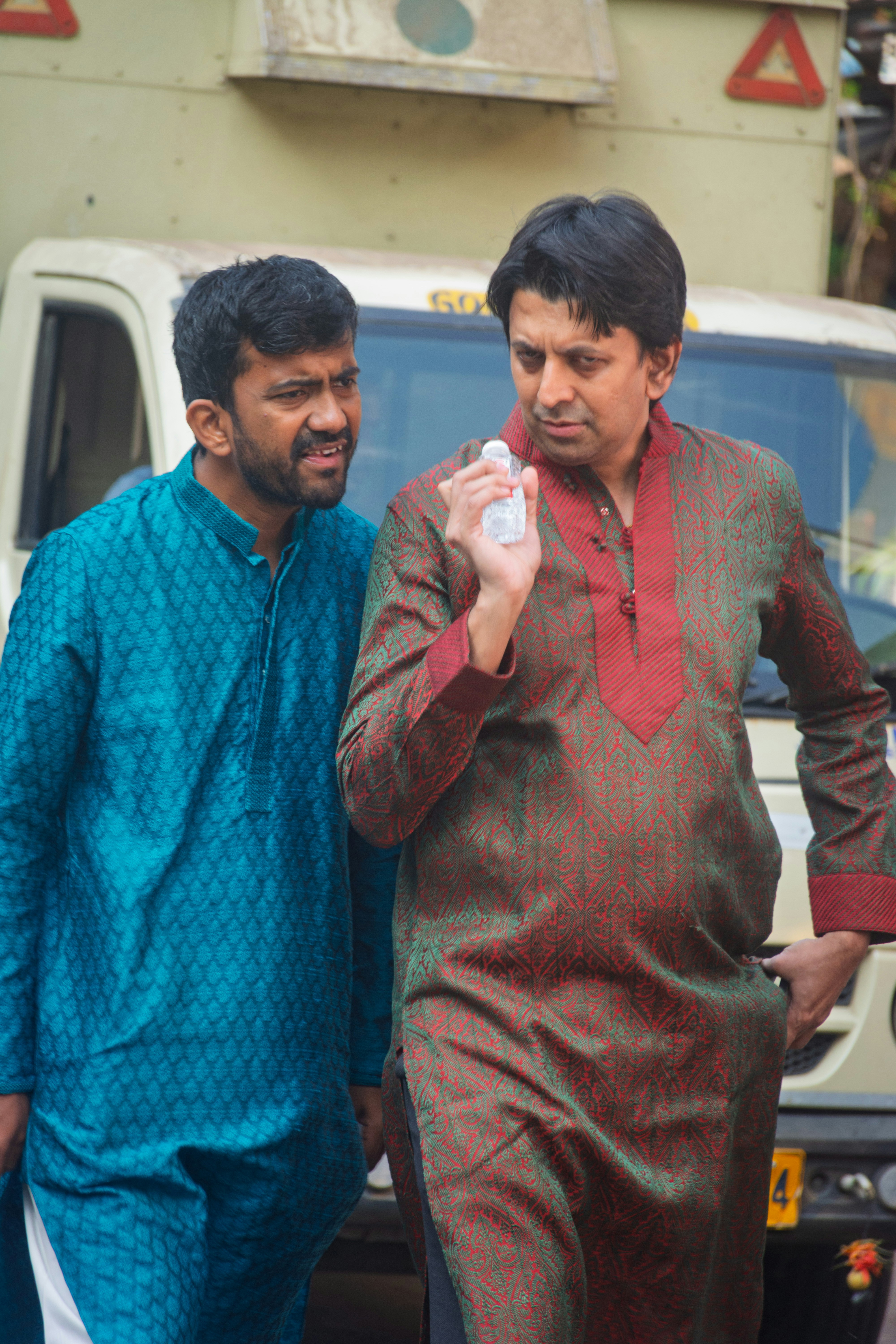 Two men in traditional attire pause beside a truck, one examining a bottle