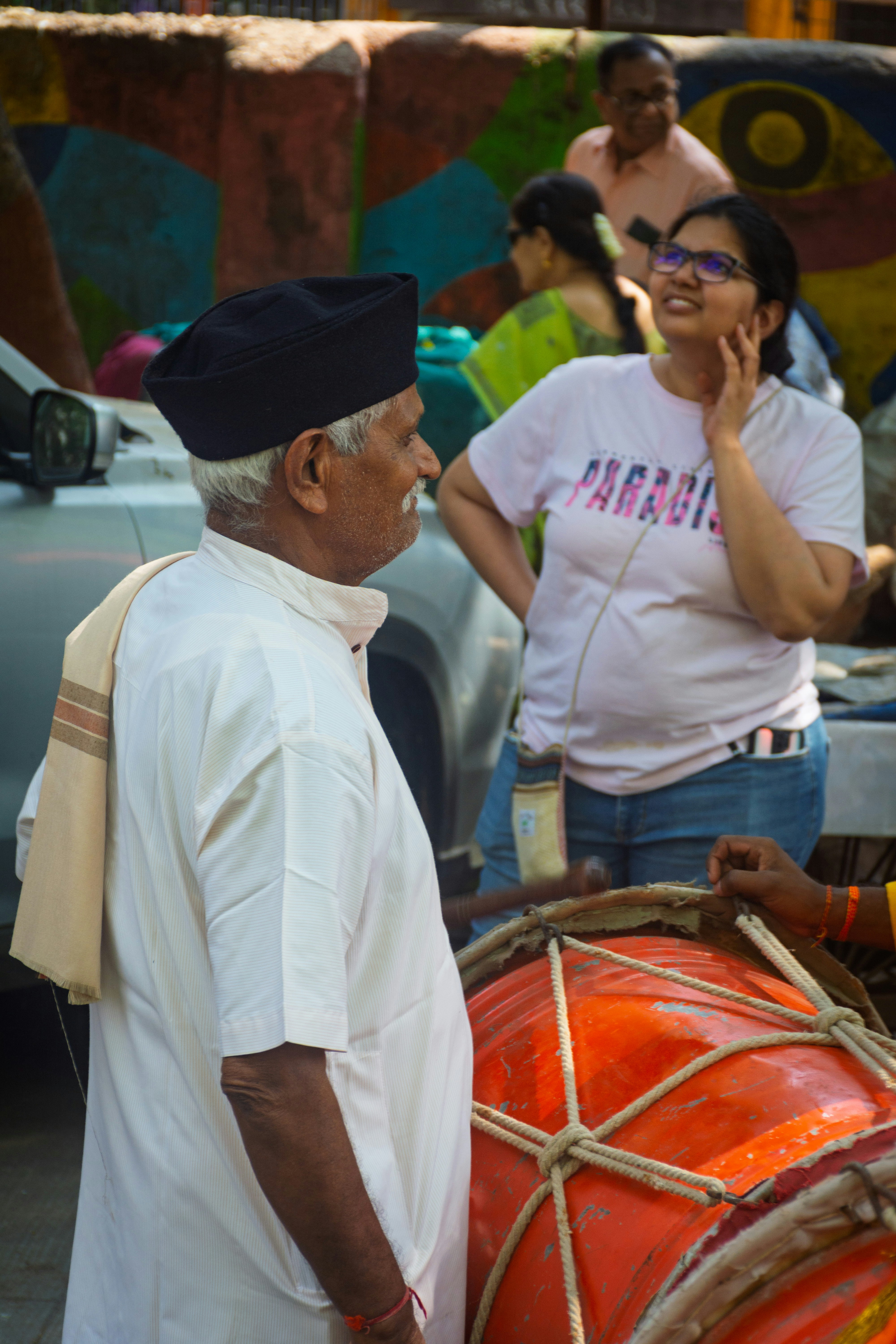 An Elderly man in a traditional white kurta watches a drum while a curious bystander smiles nearby