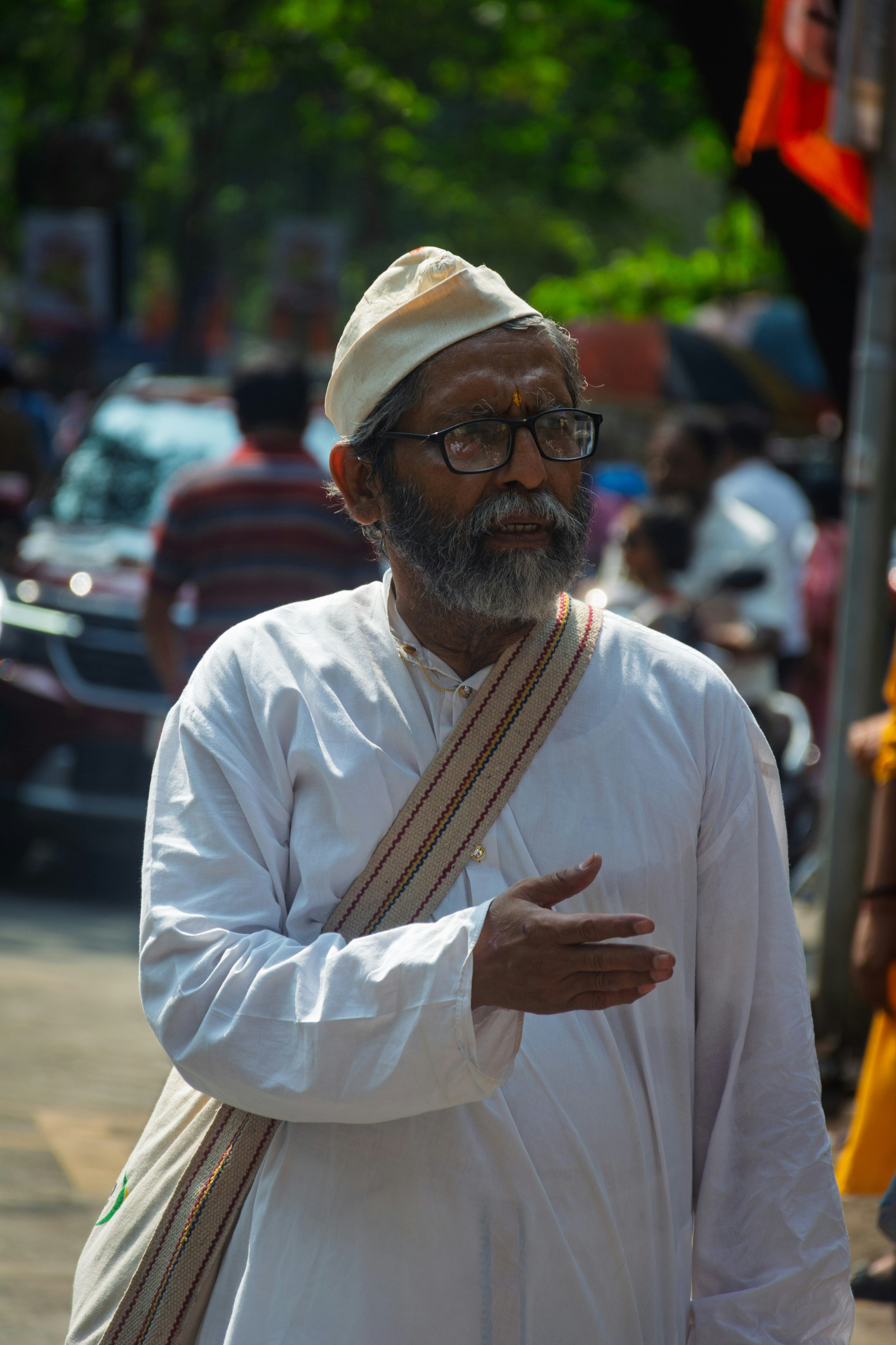 A bearded man in white traditional attire and a cream headscarf steps forward in a crowded street, carrying a woven sash. The scene emphasizes cultural attire and a moment of calm amid background activity.