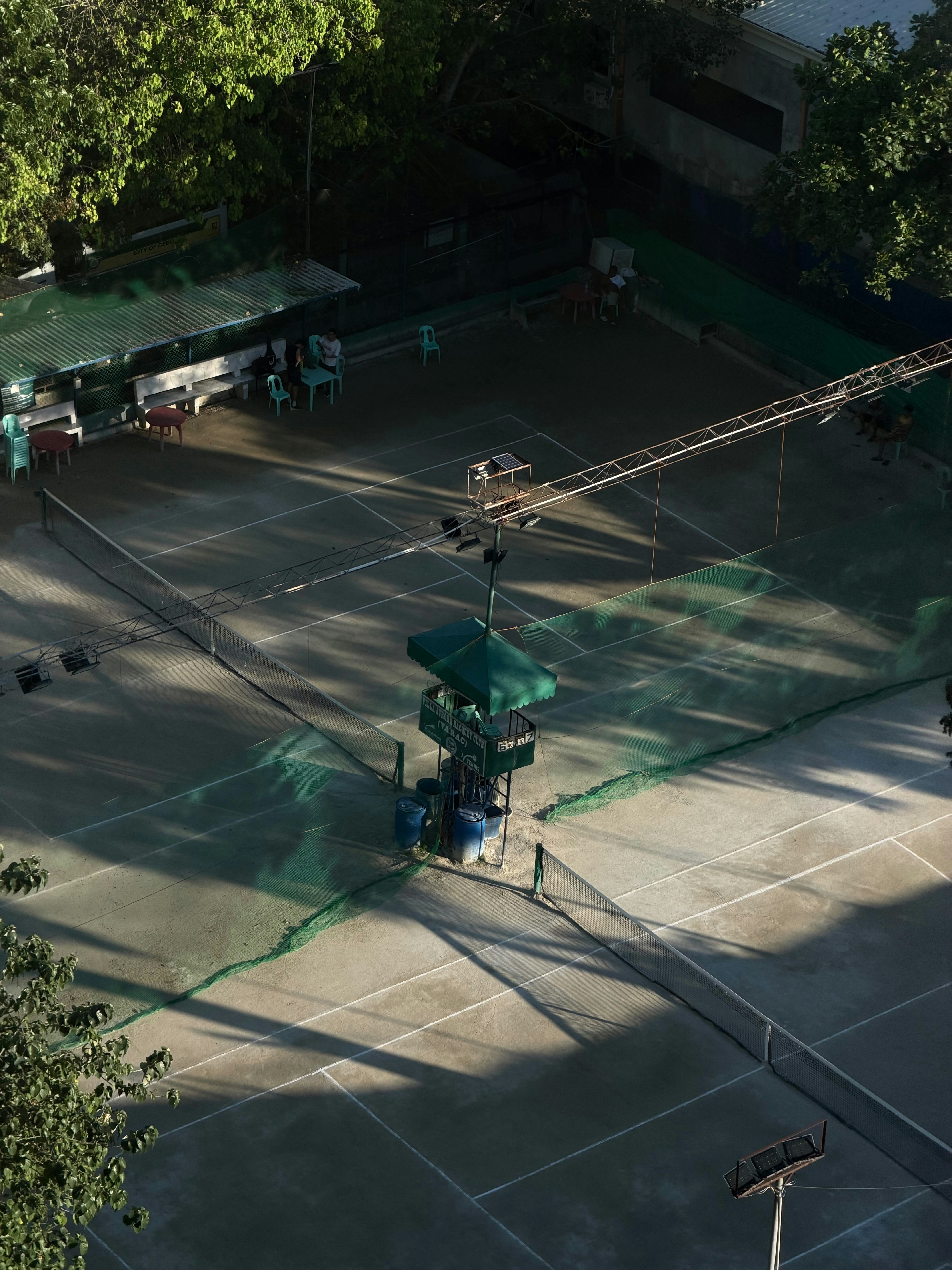 Empty tennis court under dappled shade and a lone umpire’s tower