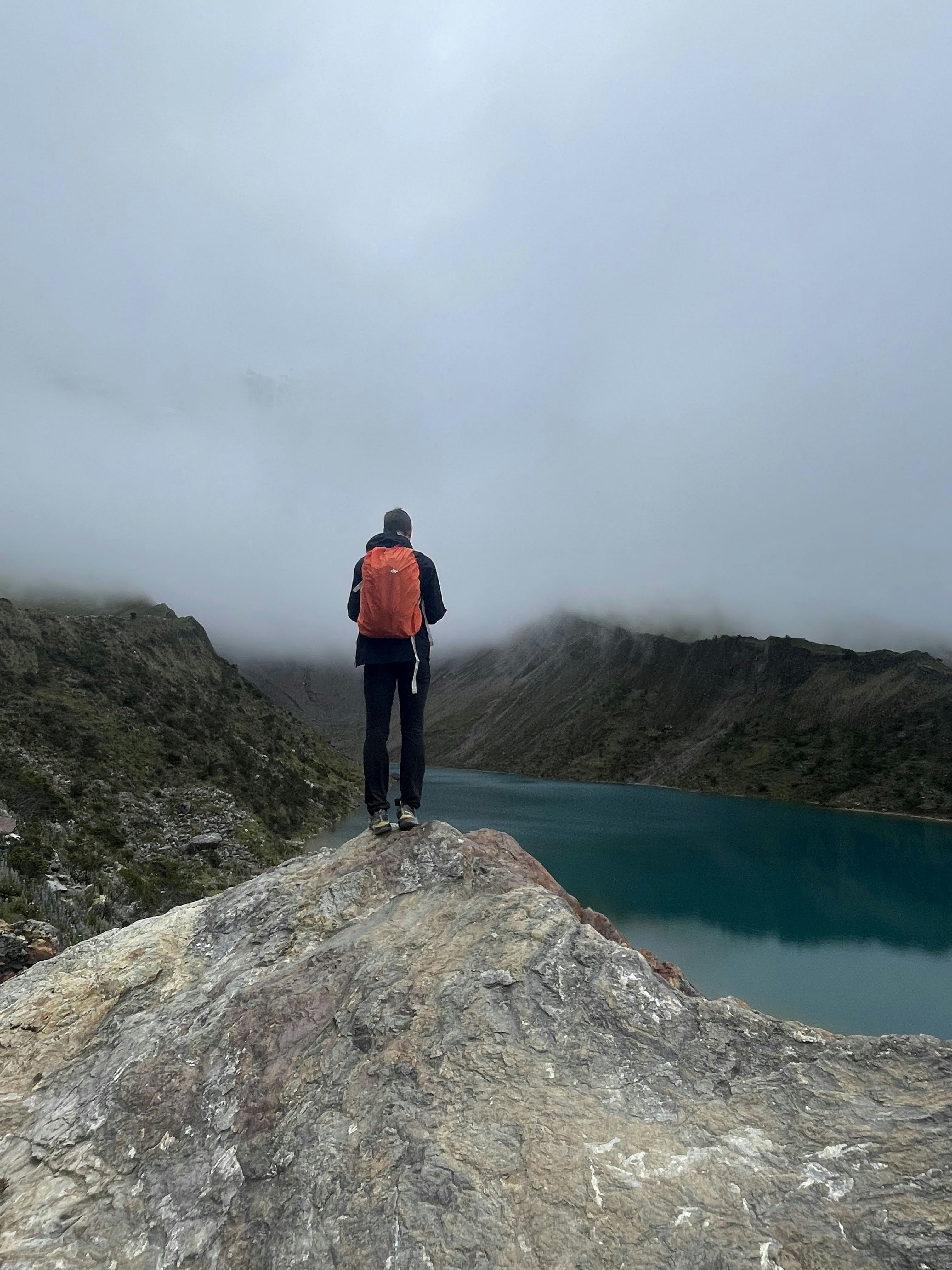 Red-Backpack Ascent: A lone hiker on a cliff overlooking a turquoise crater lake