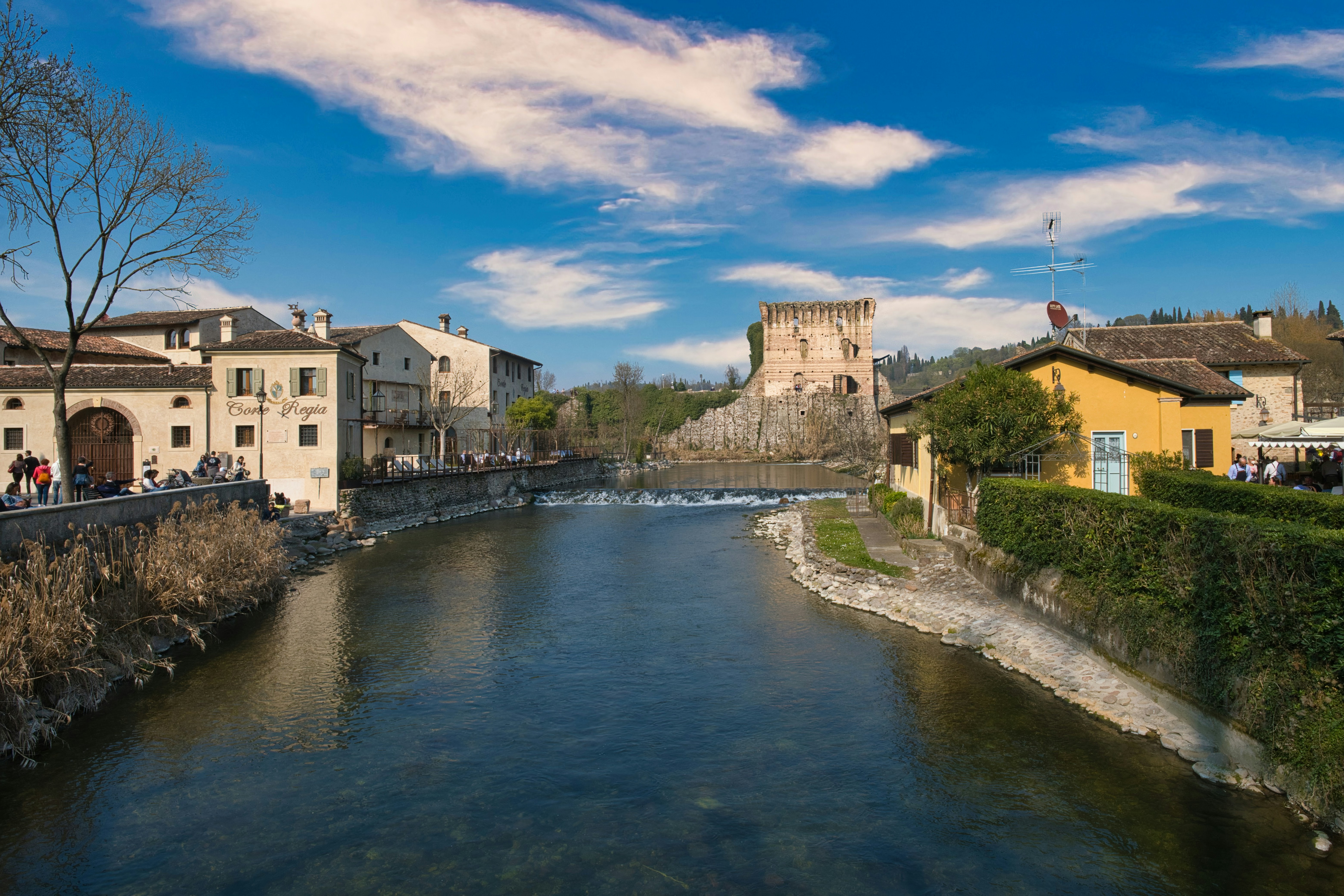 Riverside Gate: A sunlit Italian Town Framed by Historic Stone and a Swaying Castle Chimney