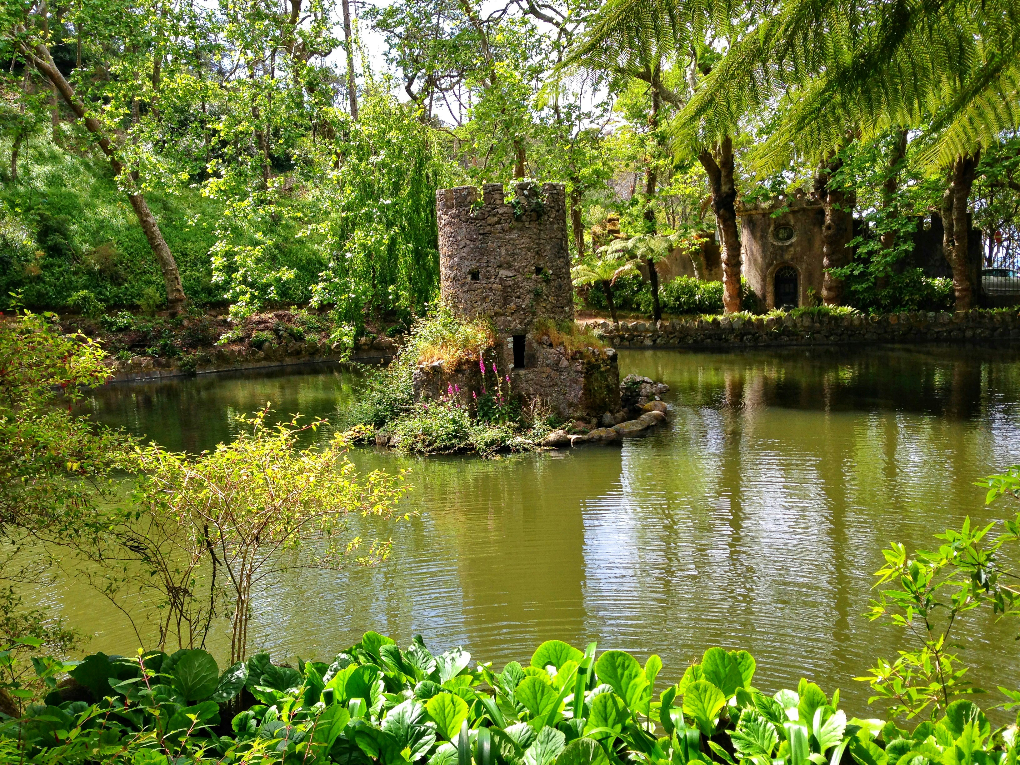 A serene garden pond features a small stone turret rising from the water, surrounded by green foliage and reflected trees. The scene emphasizes tranquil water and historic structure.