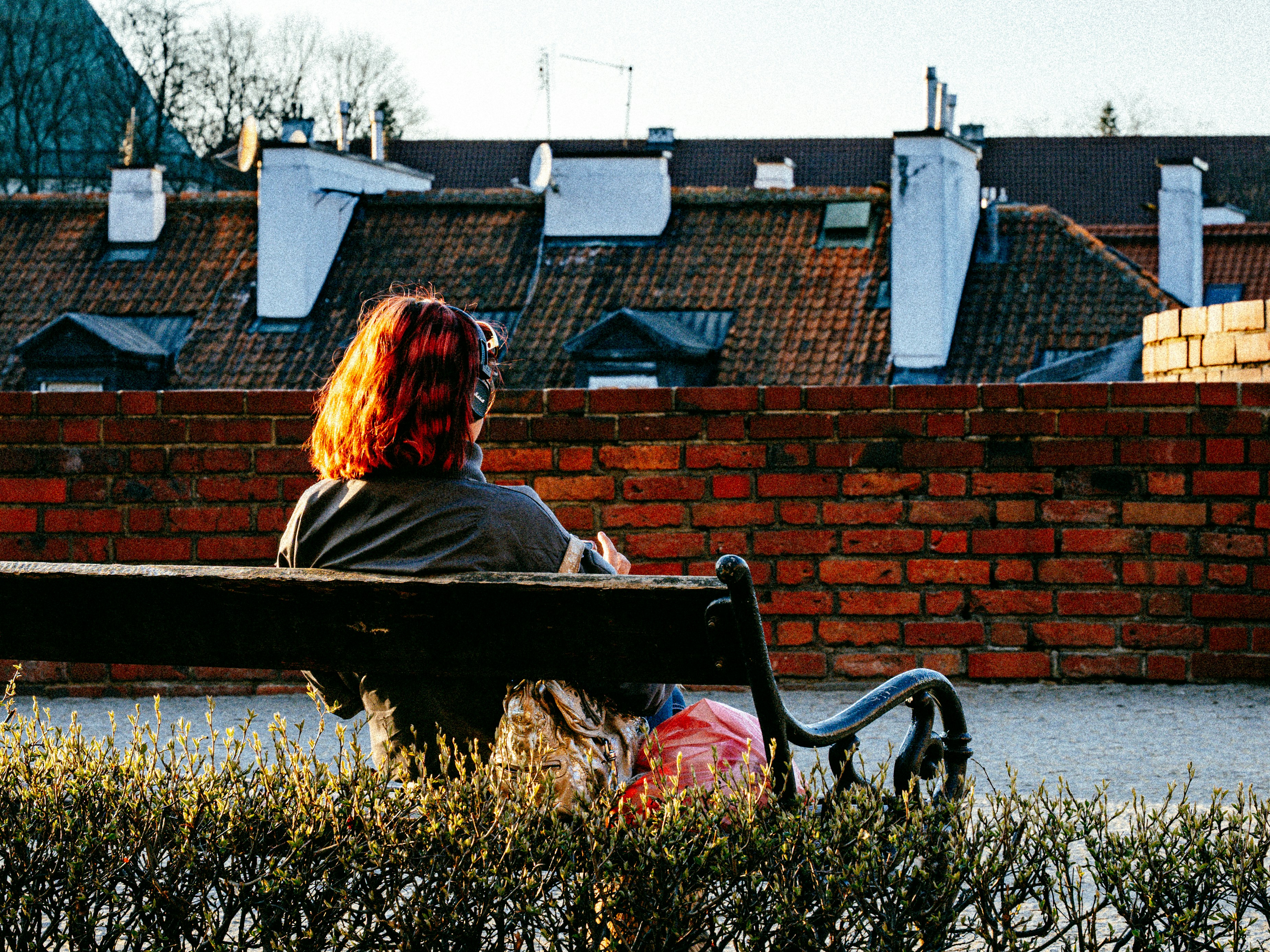 Backlit Quiet: A Woman with Red Hair on a Park Bench Overlooking a Brick Wall