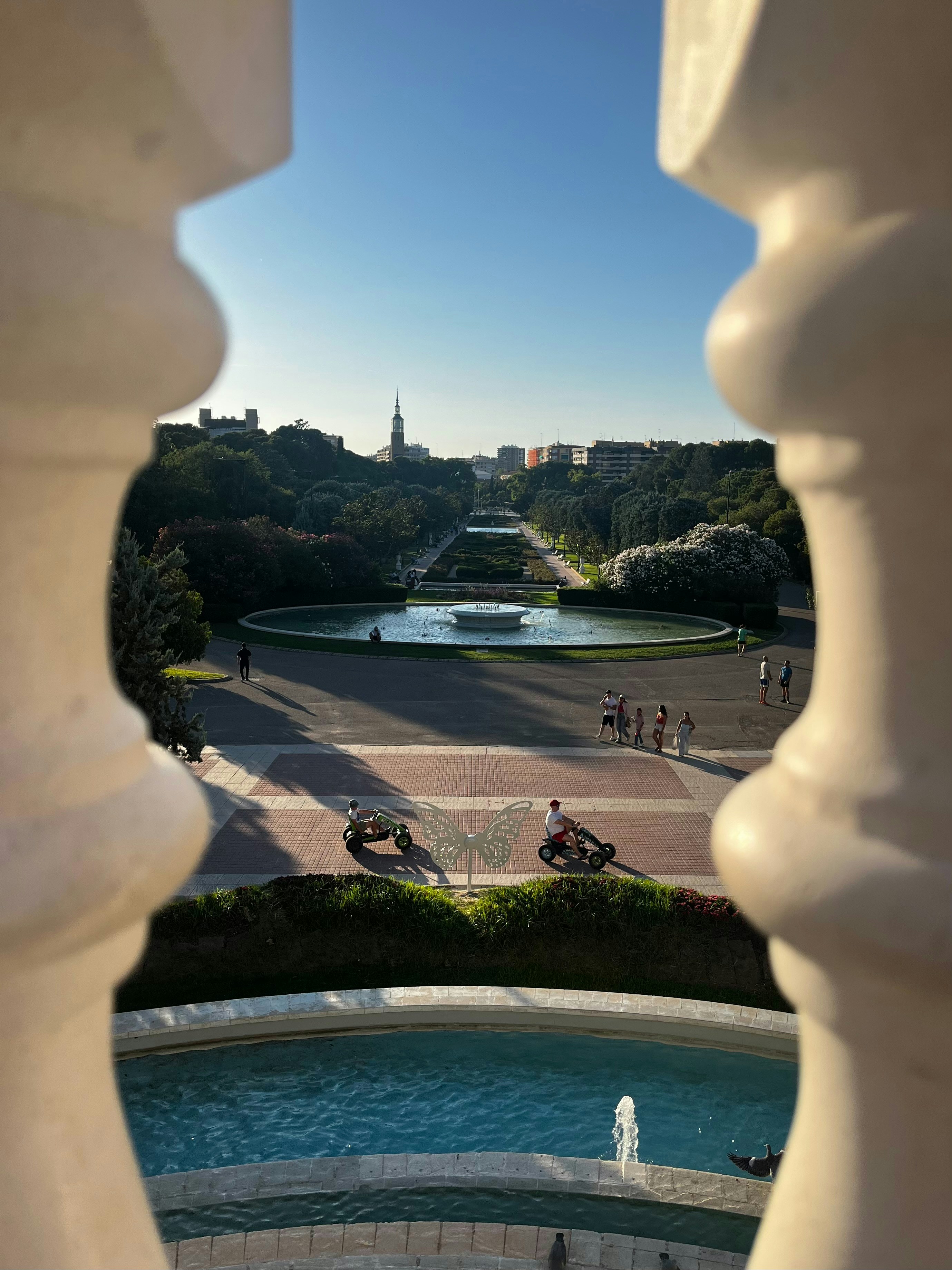 A scenic city garden vista framed by decorative balcony balustrades, with a central fountain, manicured lawns, and pathway leading toward distant skyline.