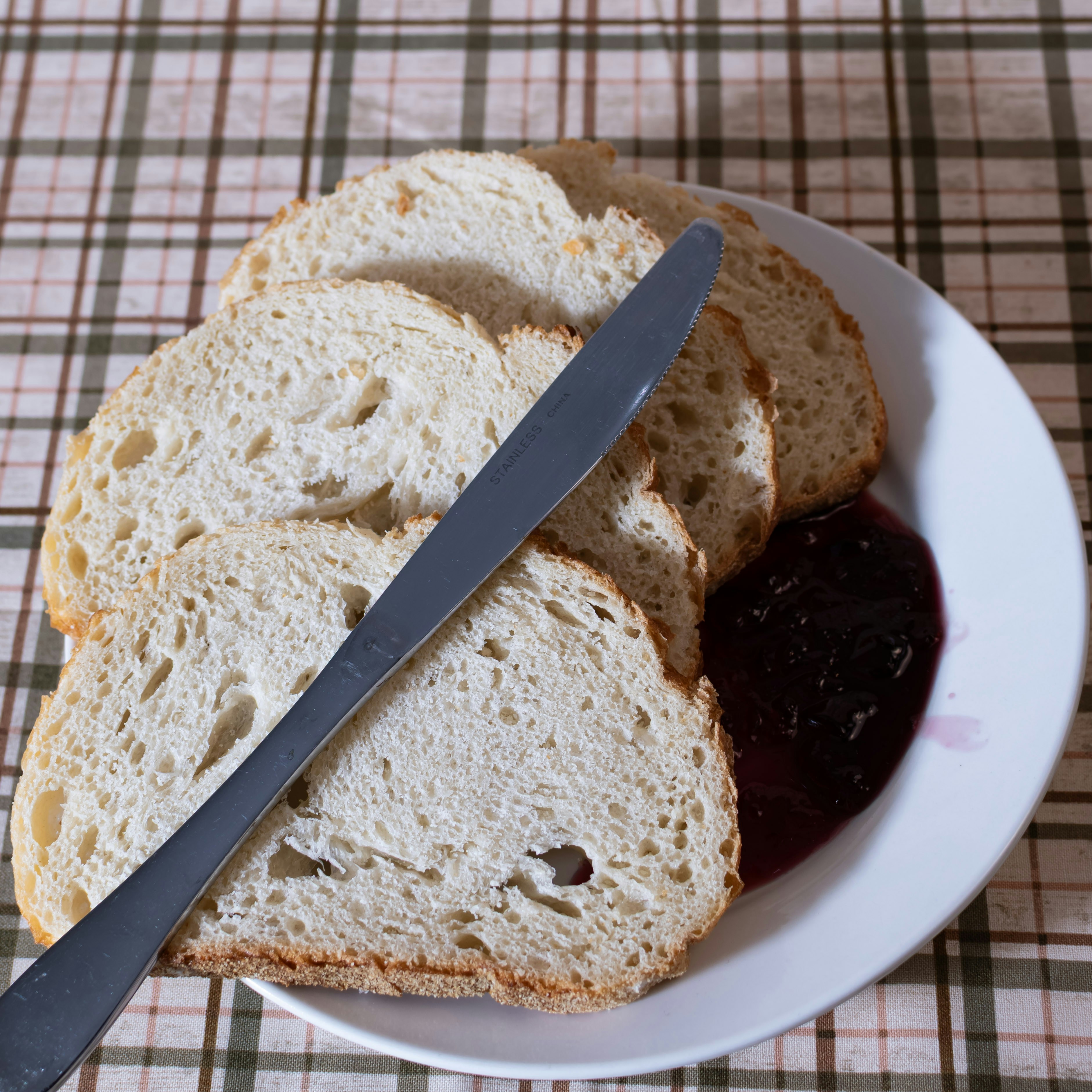 Plain white bread slices on a white plate with a smear of jam, a knife resting across the loaf, on a plaid tablecloth.