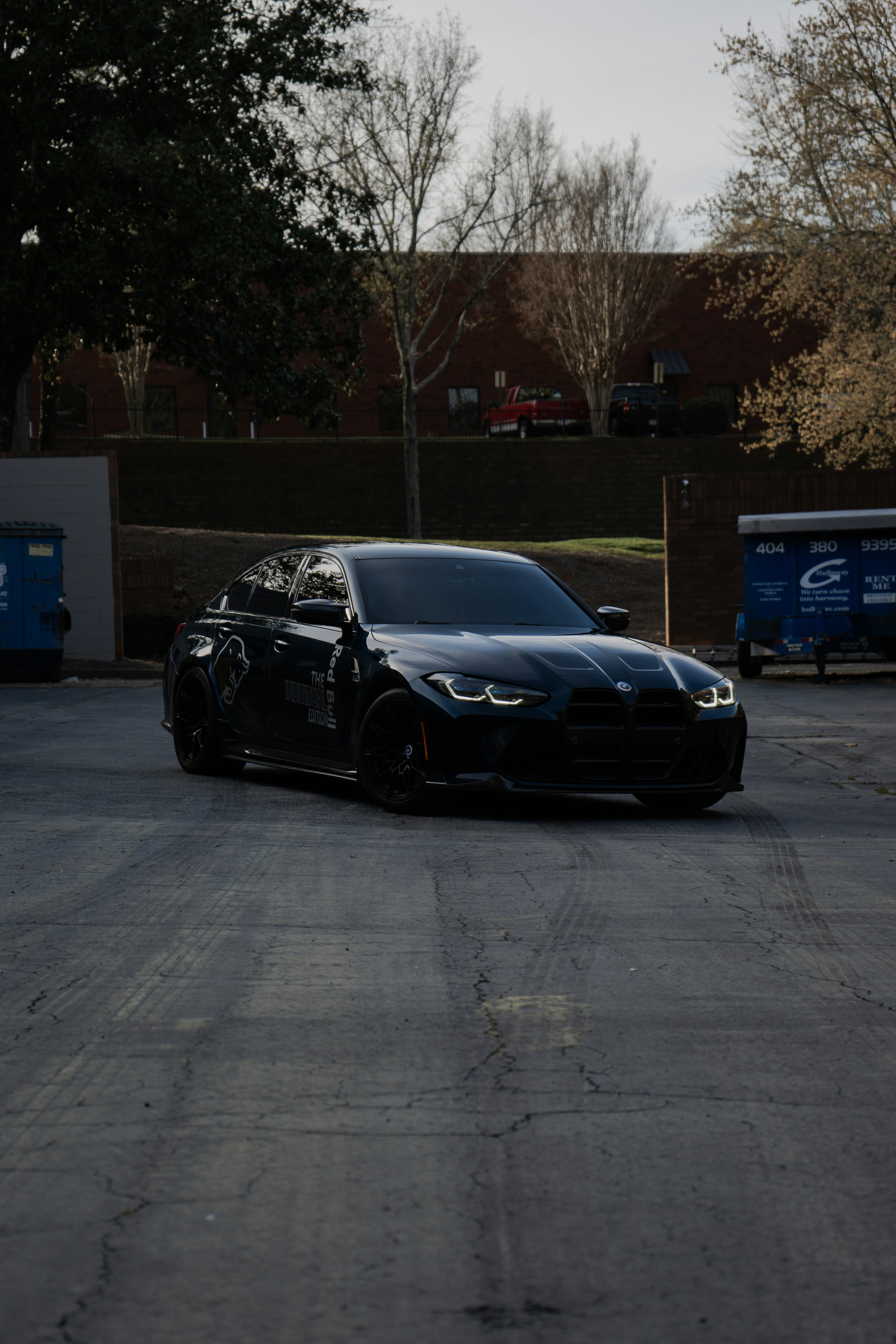 Sleek black car sits centered in a quiet parking lot with subdued background trees and dumpsters; a low-contrast, moody street scene highlighting the vehicle's silhouette.