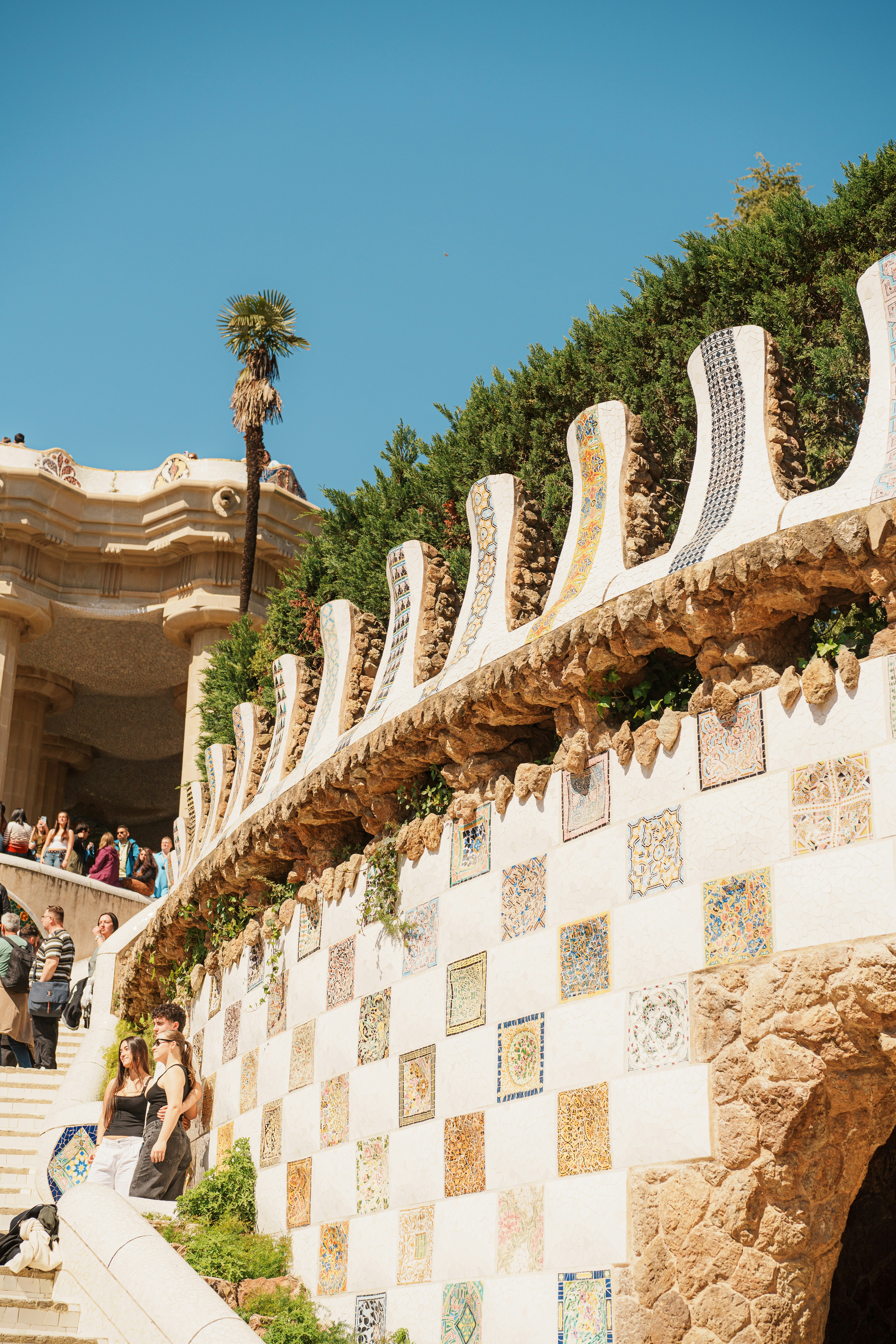 Colorful tiled wall and organic stone railing along a sunlit terrace at Parc Güell, with a few visitors descending steps beside a lush hedge and a tall palm against a clear blue sky.