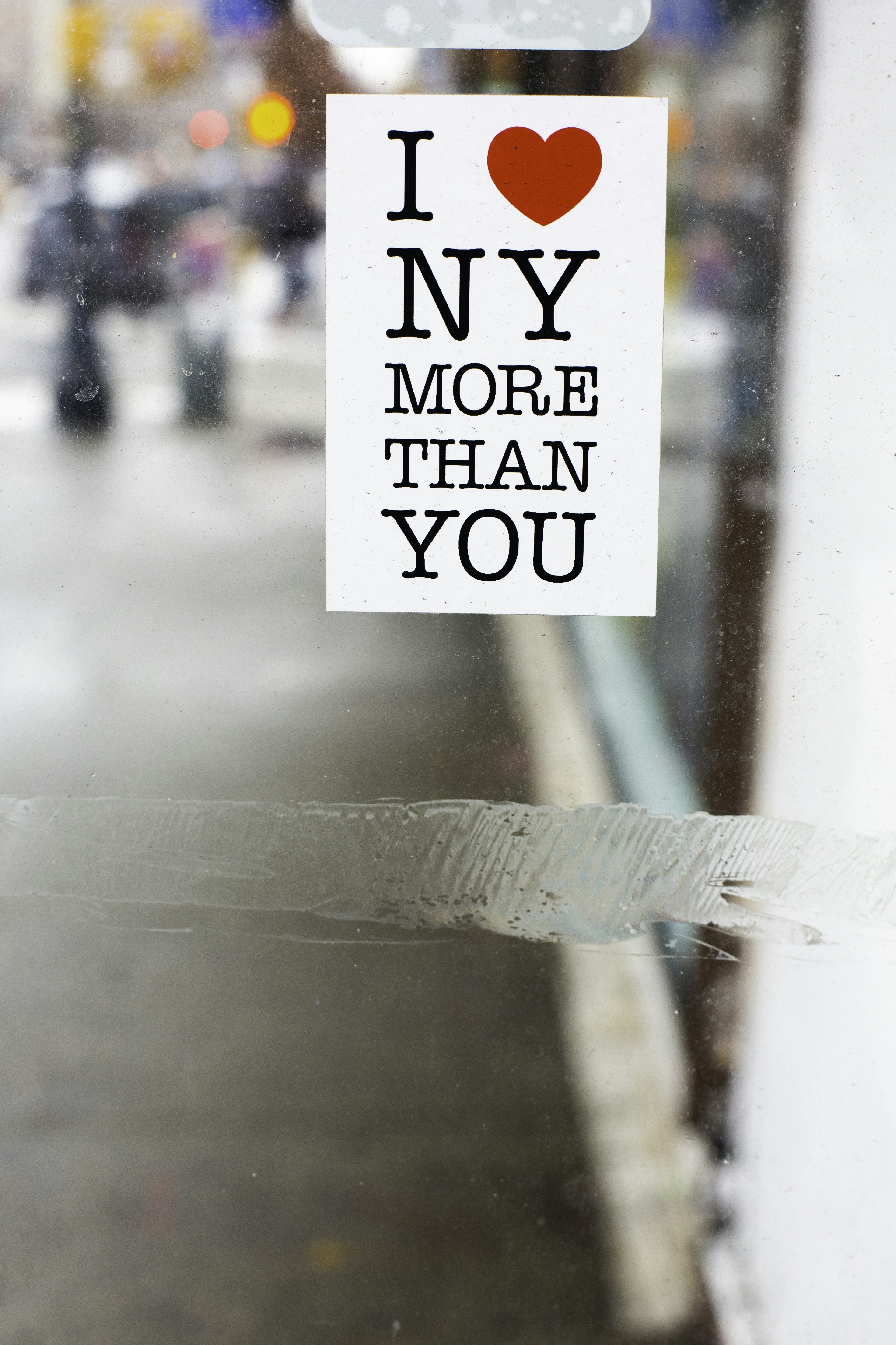 A white sticker on a grimy window reads “I ♥ NY MORE THAN YOU,” with soft city lights and pedestrians blurred in the background.