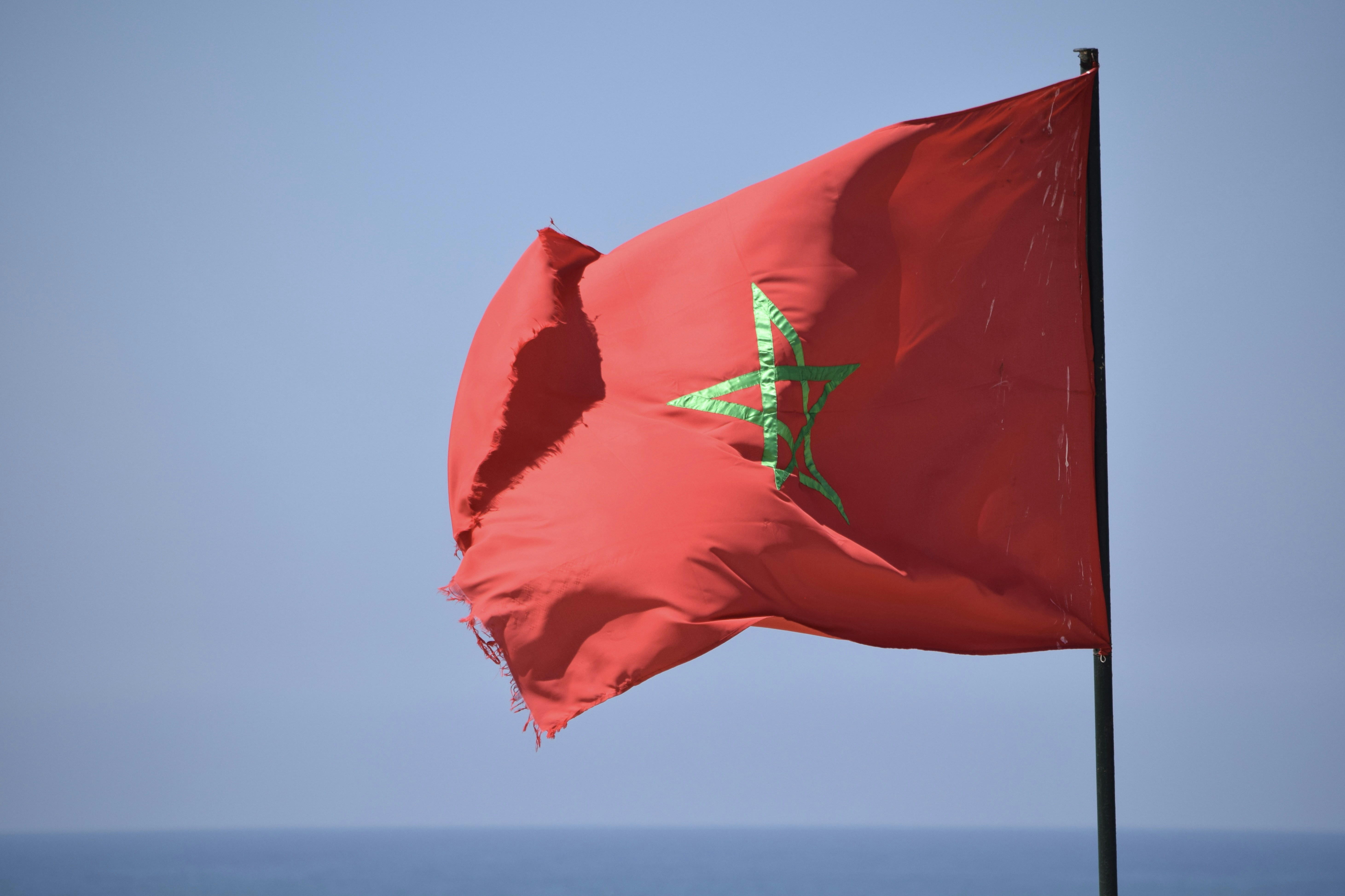 Flag of Morocco: wind-swept crimson banner with green star against a calm sea horizon