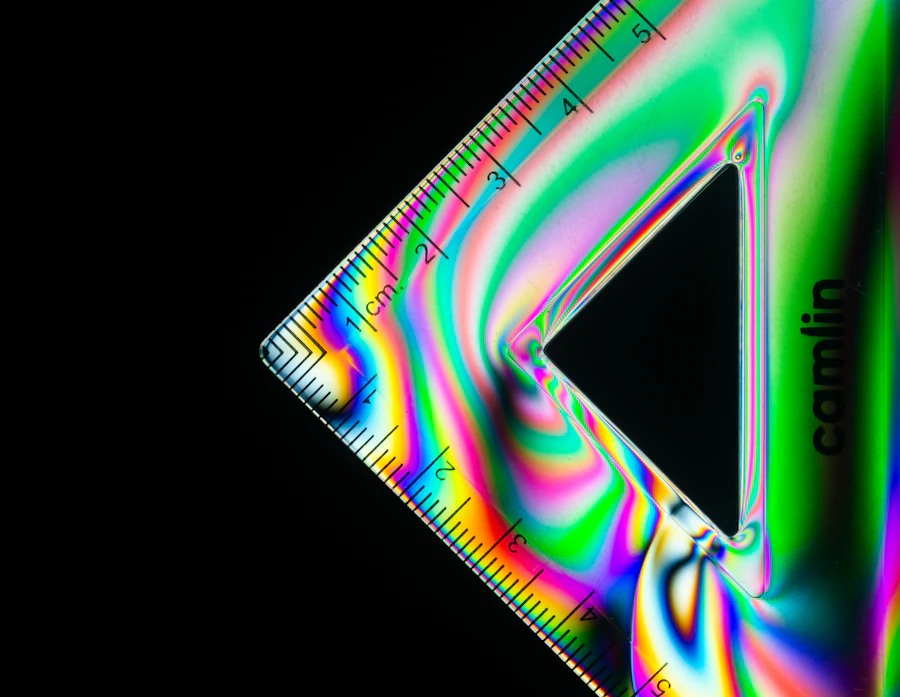 Close-up of a colorful triangular ruler against a black background, revealing vivid iridescent hues and precise markings. This is a macro-style study focused on the tool’s edge geometry and reflective surface.