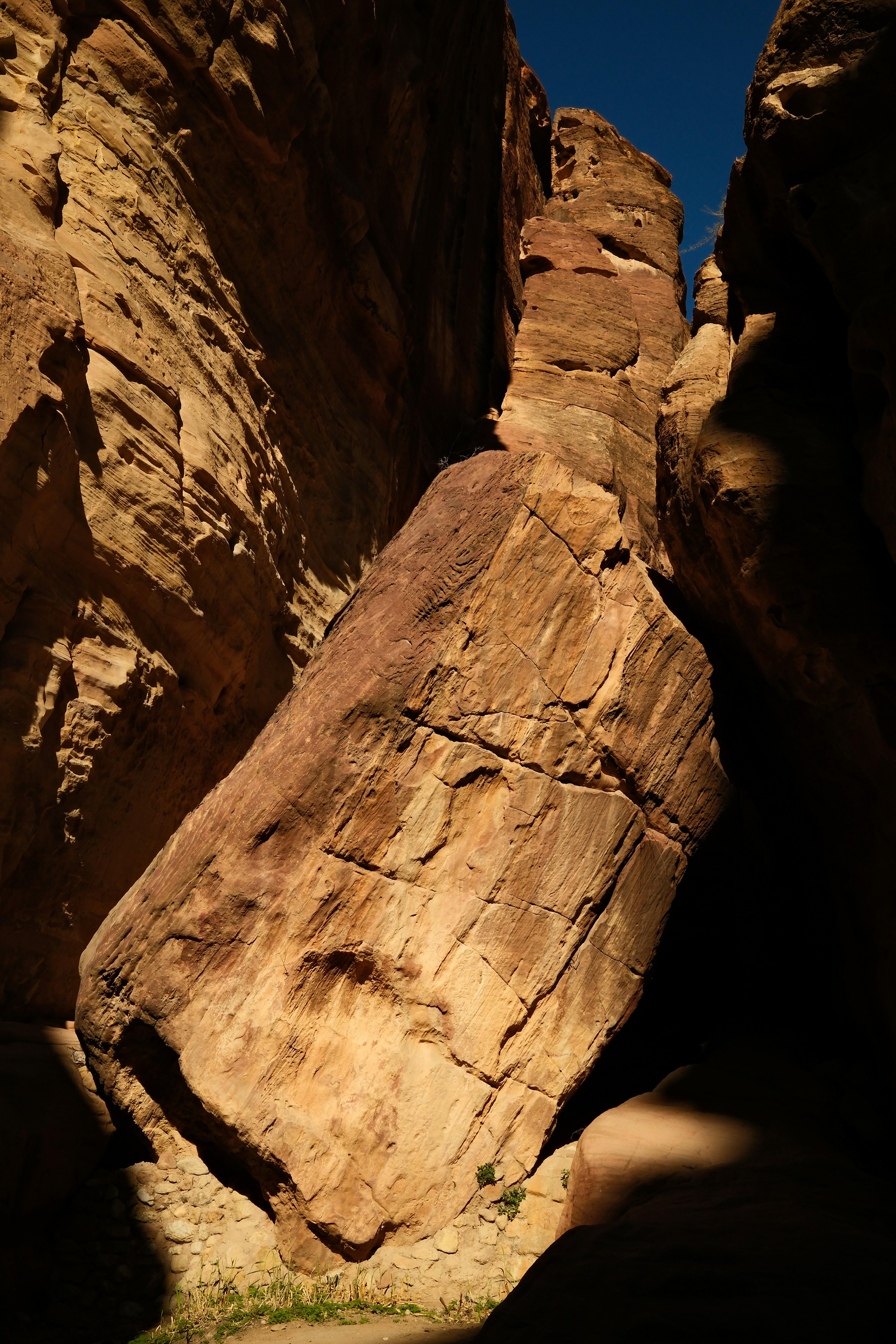A dramatic close-up of a massive, fractured sandstone boulder wedged precariously within a narrow gorge in Petra's Siq. Direct sunlight hits the precariously balanced rock, highlighting its deep cracks and gritty texture, while the surrounding canyon walls remain in deep, imposing shadow, emphasizing a tense sense of monumental balance.