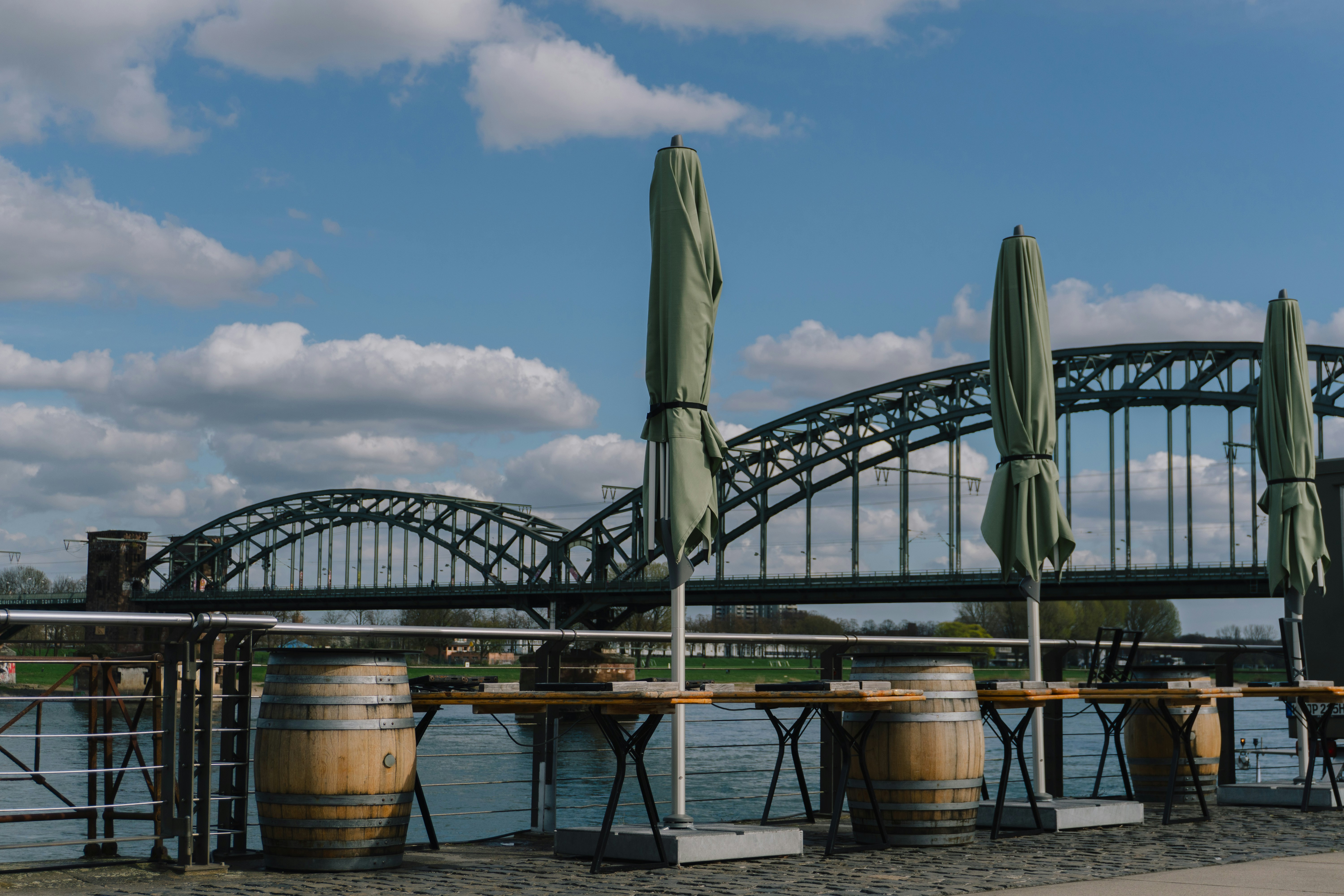 Riverfront Seating Beneath Closed Umbrellas With a Steel Arch Bridge in the Distance