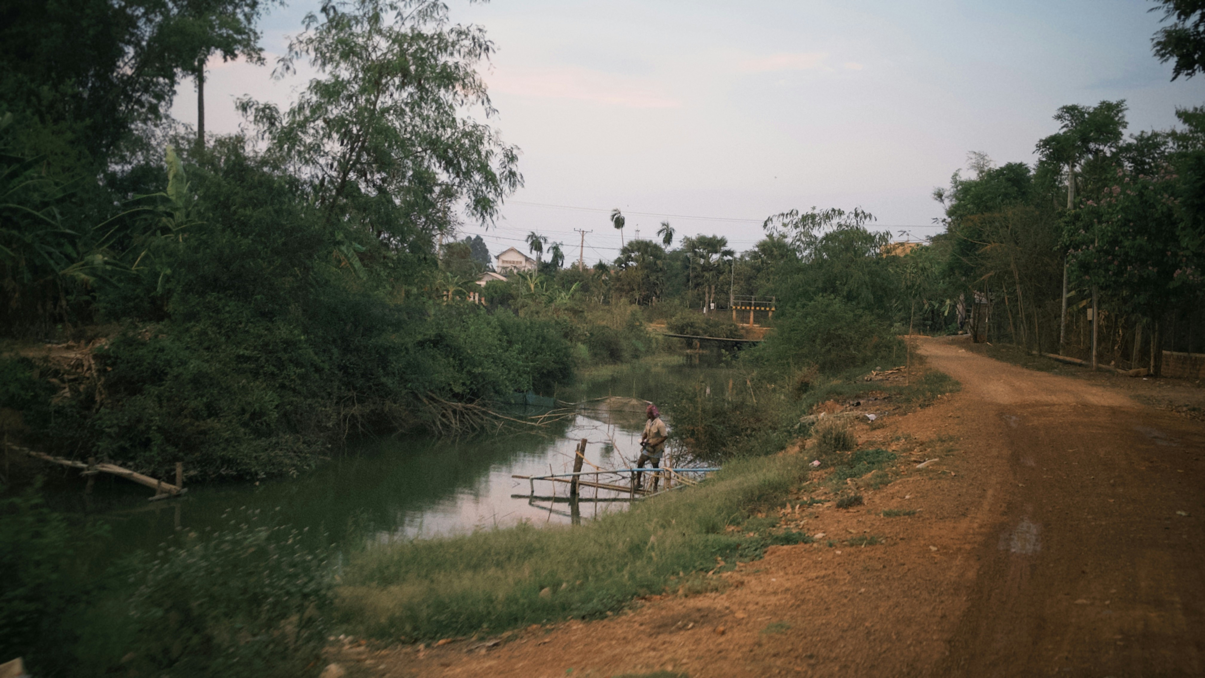 Two figures on a muddy riverbank ferry a makeshift raft along a quiet rural waterway