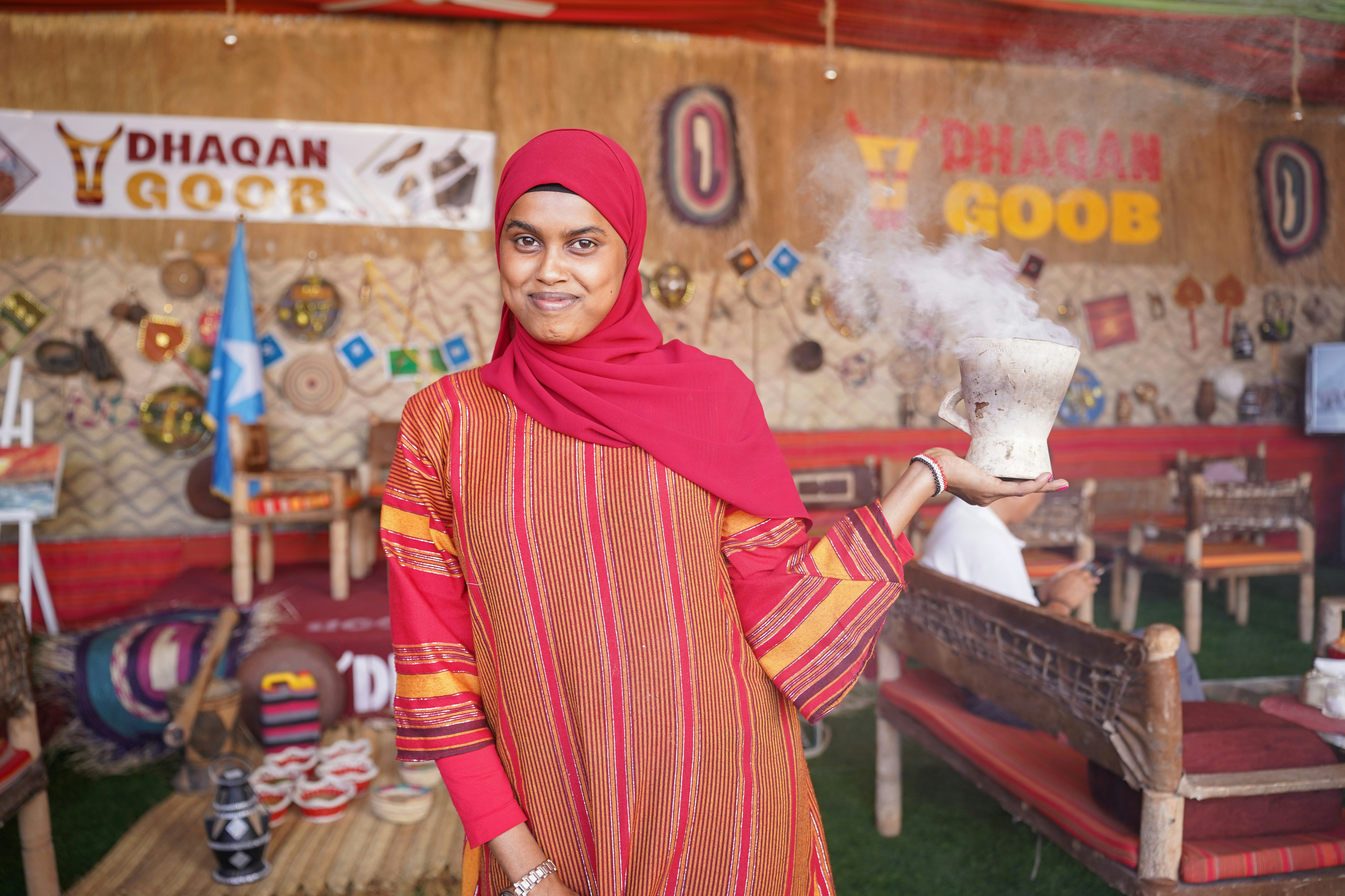 Brightly dressed woman in red hijab holds a donation bucket at a festive stall, with decorative banners and crafts in the background.