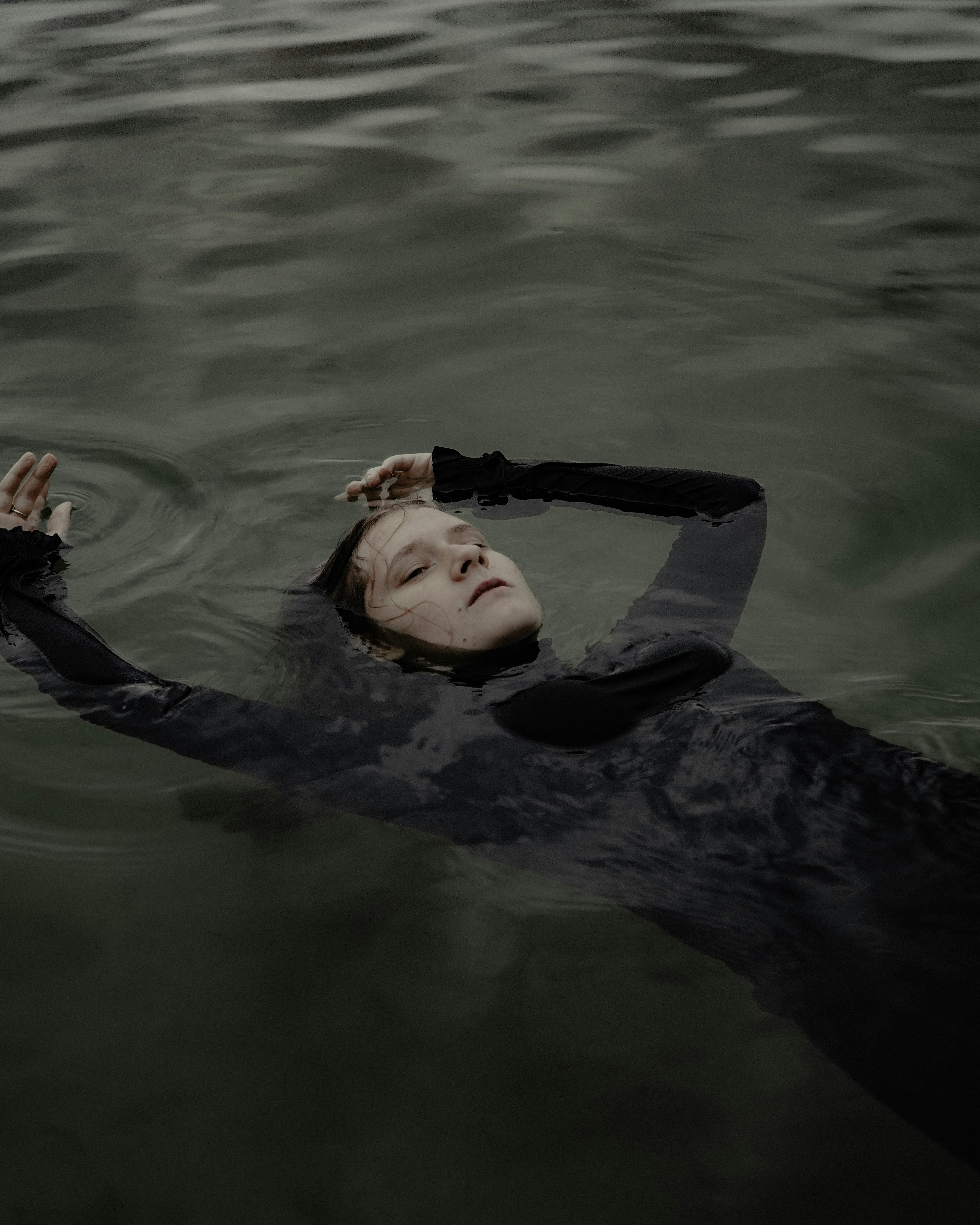 A person floats face-up in dark-green water, arms extended, wearing a black outfit; the scene centers on a serene, restrained mood without visible surroundings.