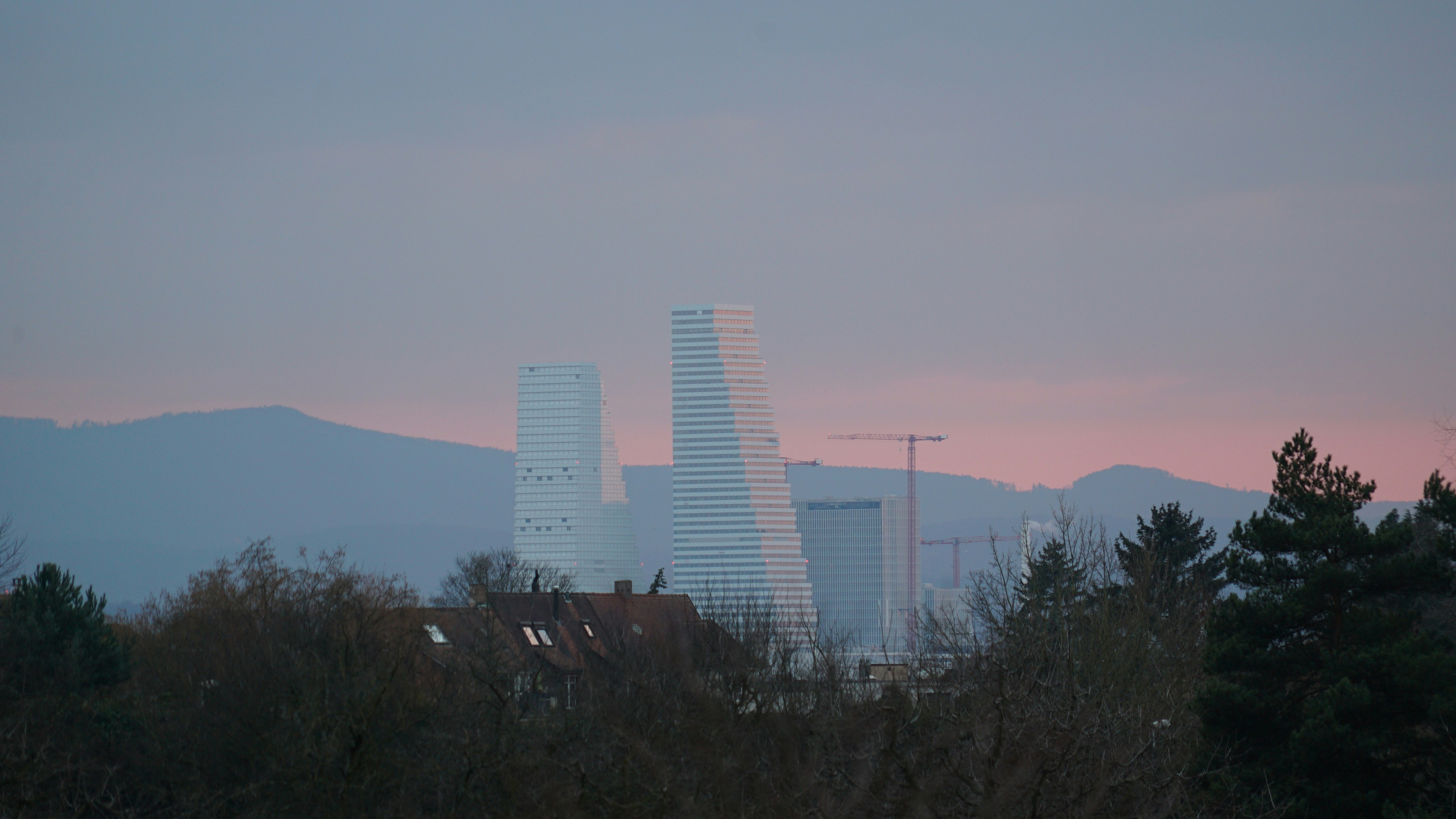 Dusk-lit city skyline rising beyond a suburban treeline; two modern tower silhouettes stand out against a pastel sky.