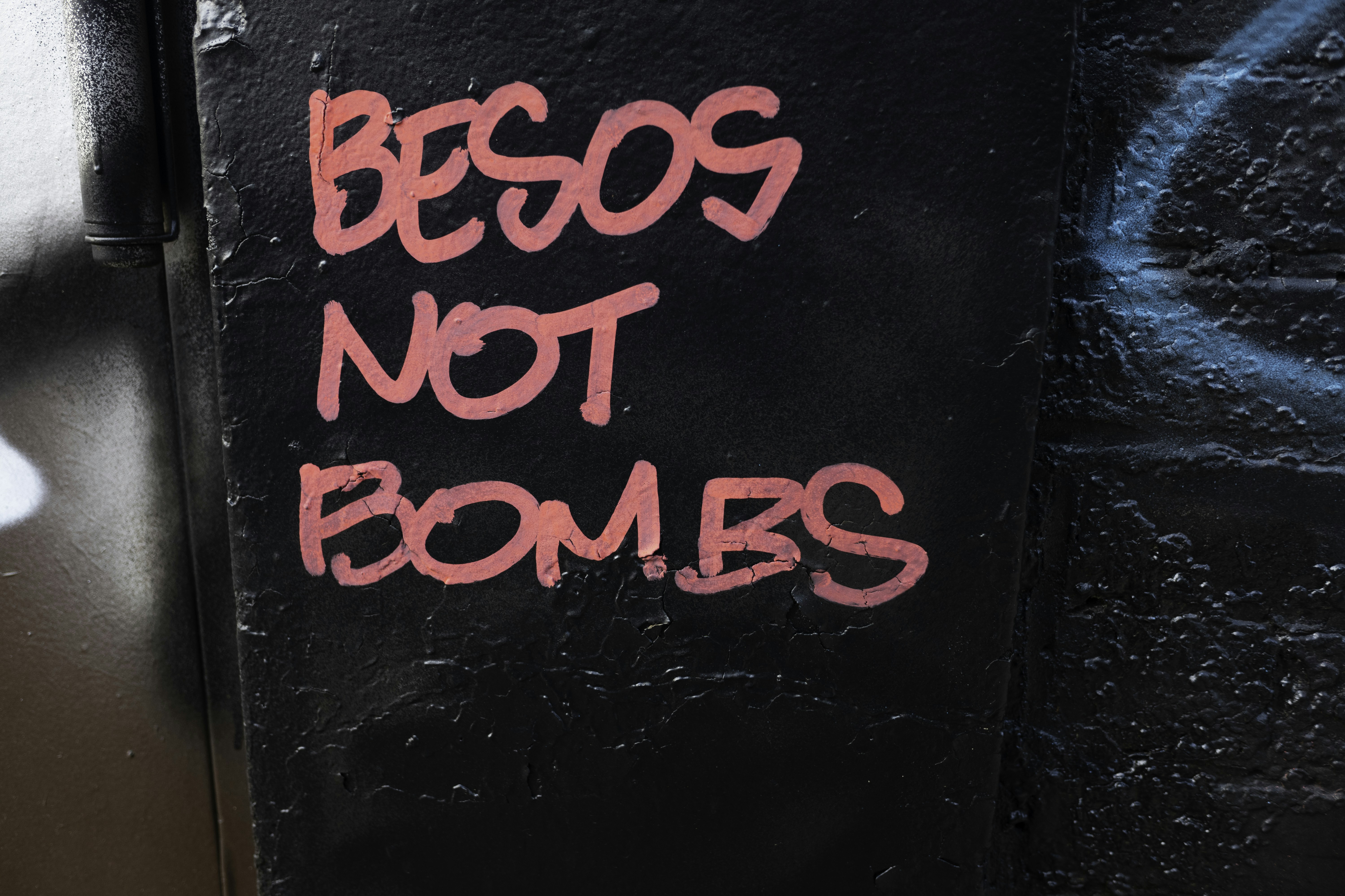 Pink graffiti reading 'BESOS NOT BOMBS' on a black, textured wall.