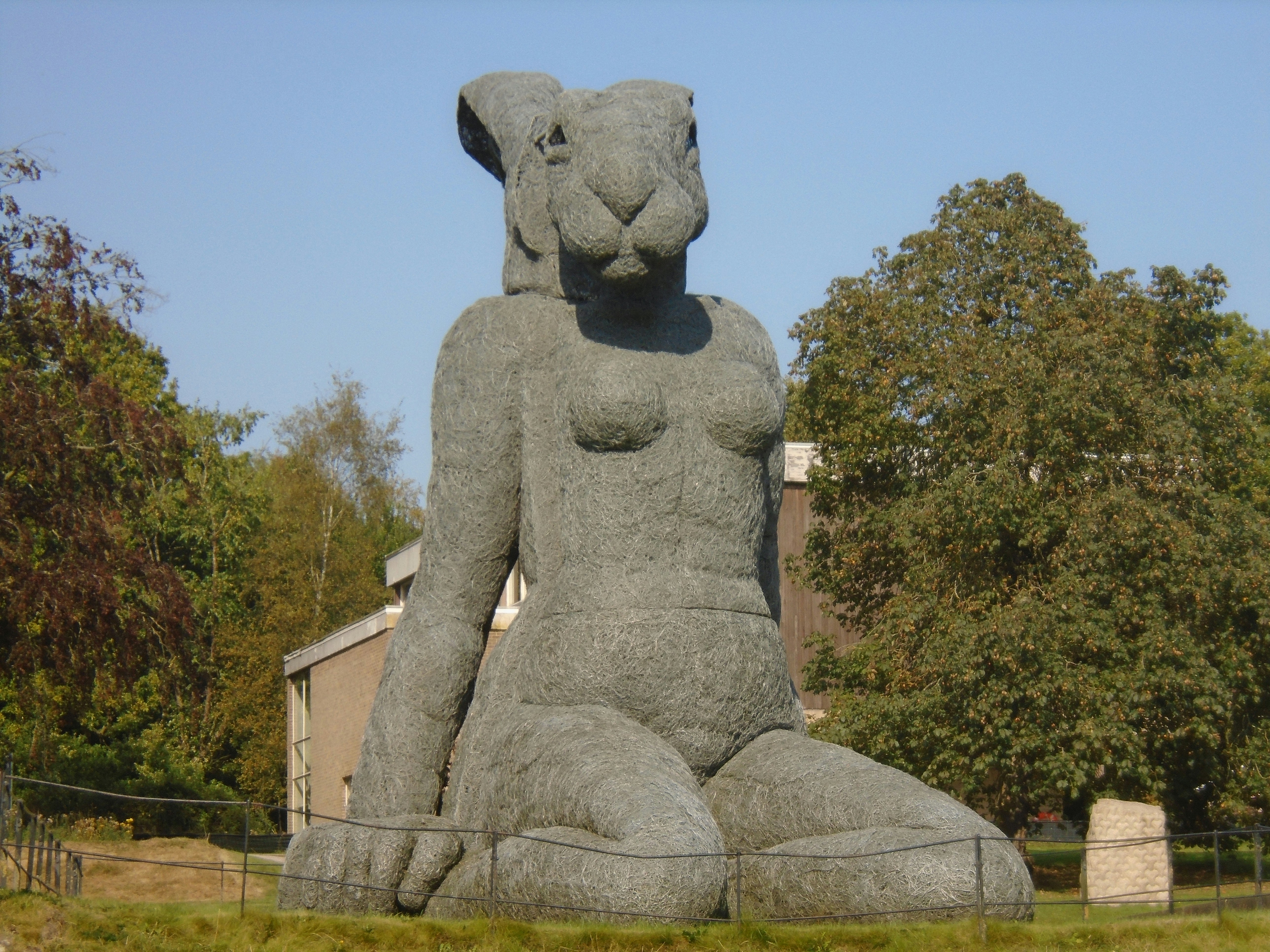 Stone panther statue seated in a park with trees and buildings in the background. A quiet, outdoor sculpture scene.