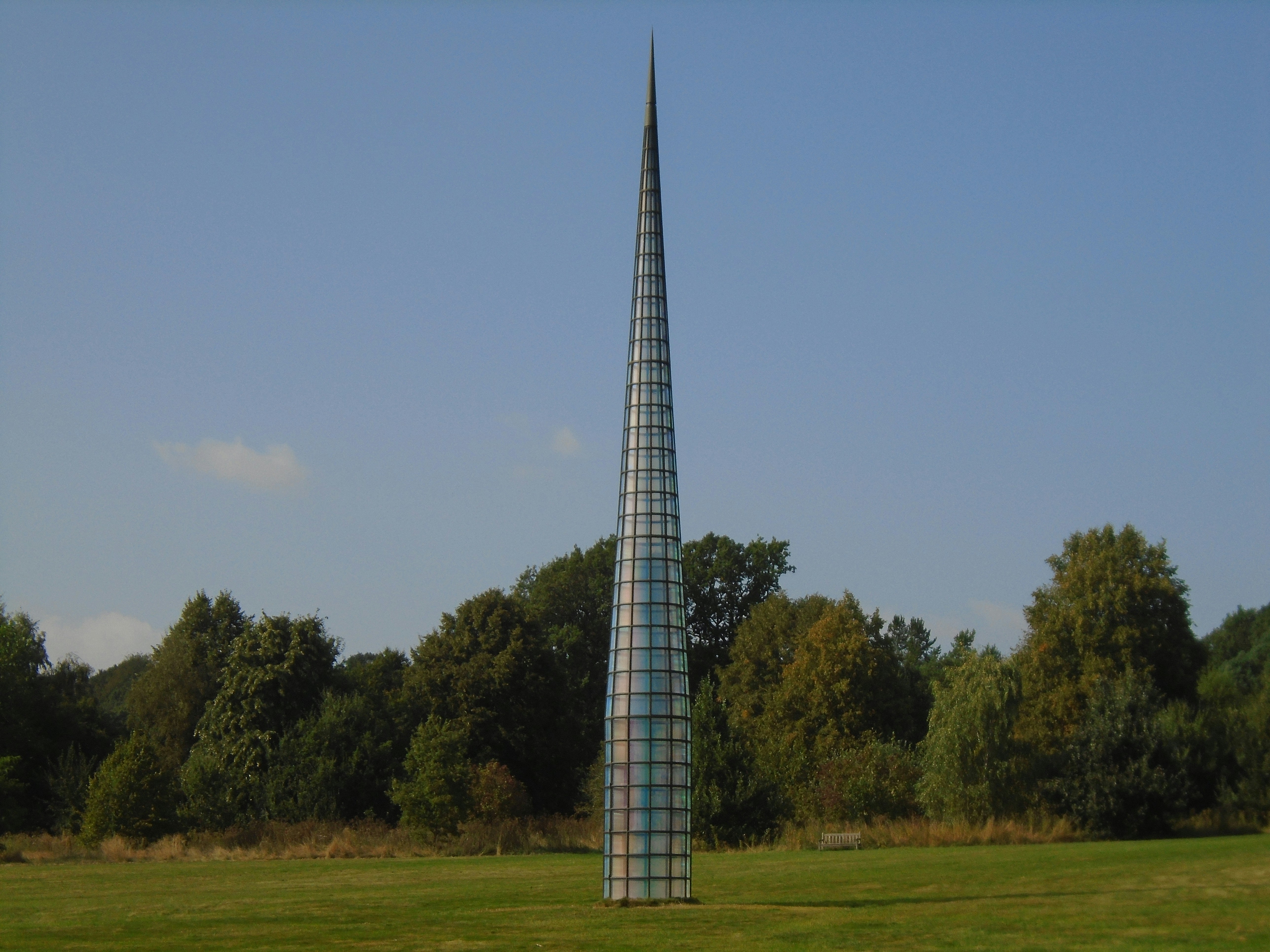 A tall, glass-covered obelisk rises from a grassy clear area, bordered by trees under a clear blue sky.