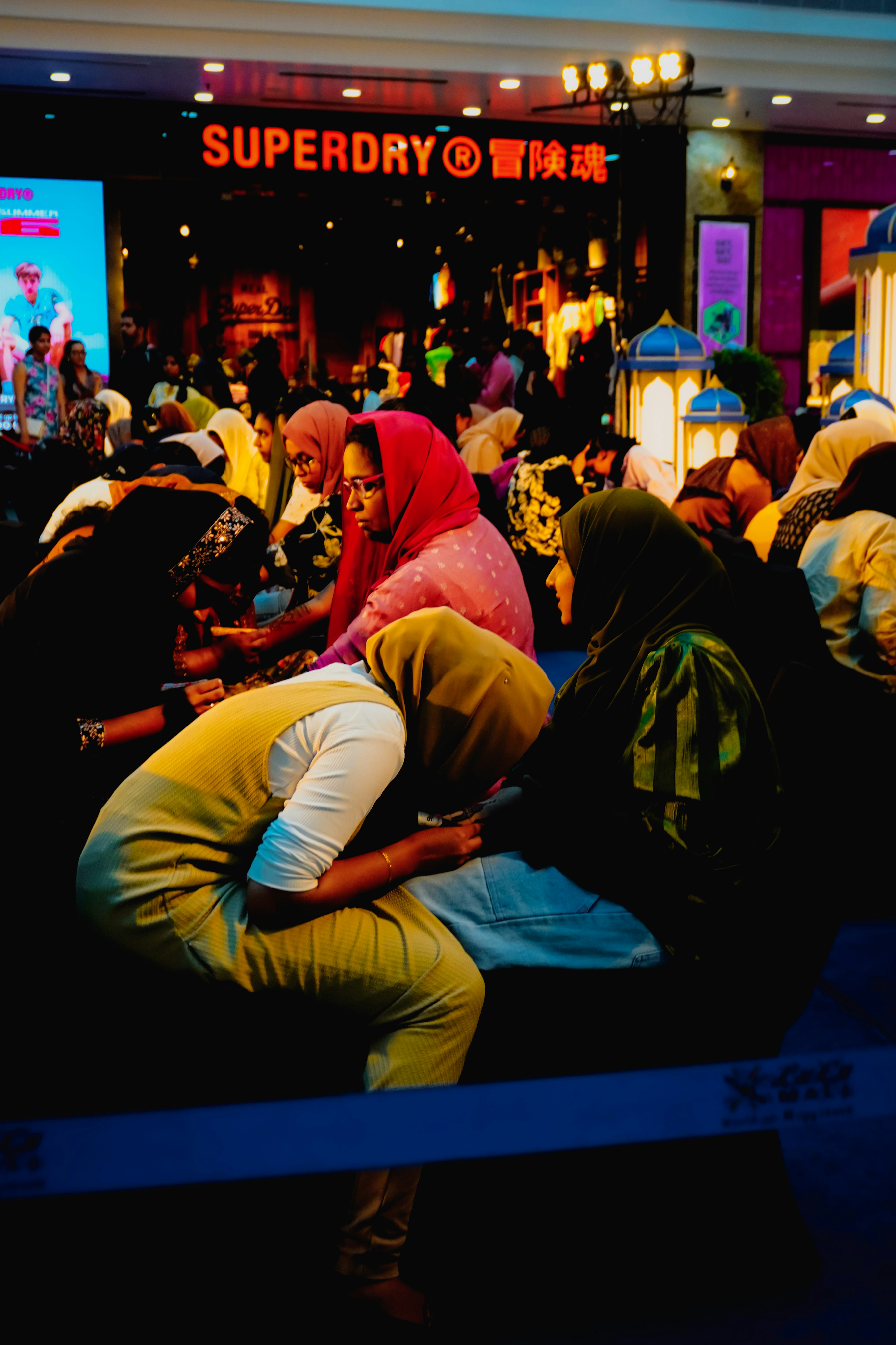 Crowd of women in hijabs gathered around a shared activity under a lit storefront