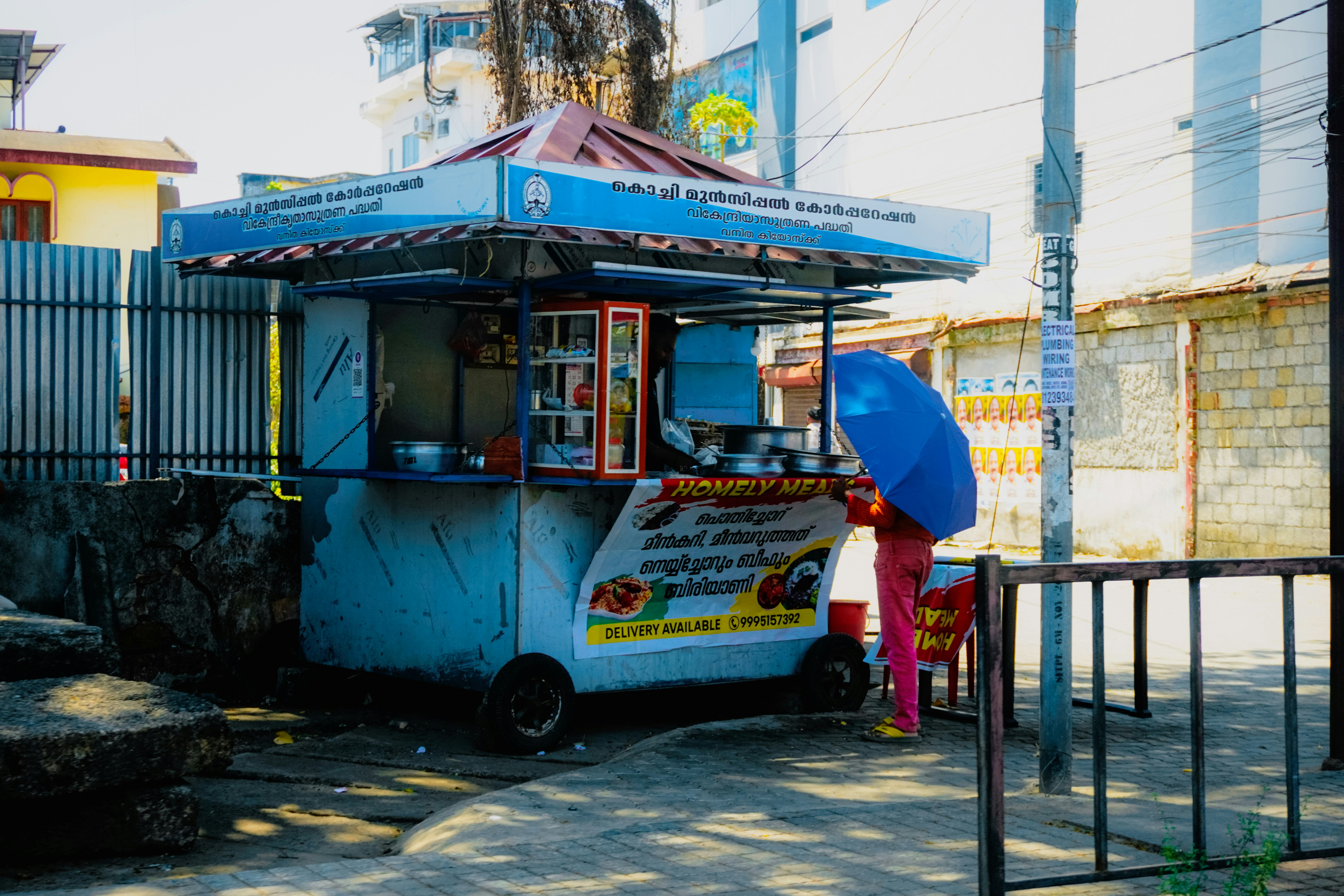 Blue street food cart under a canopy, with a person in pink beneath an umbrella