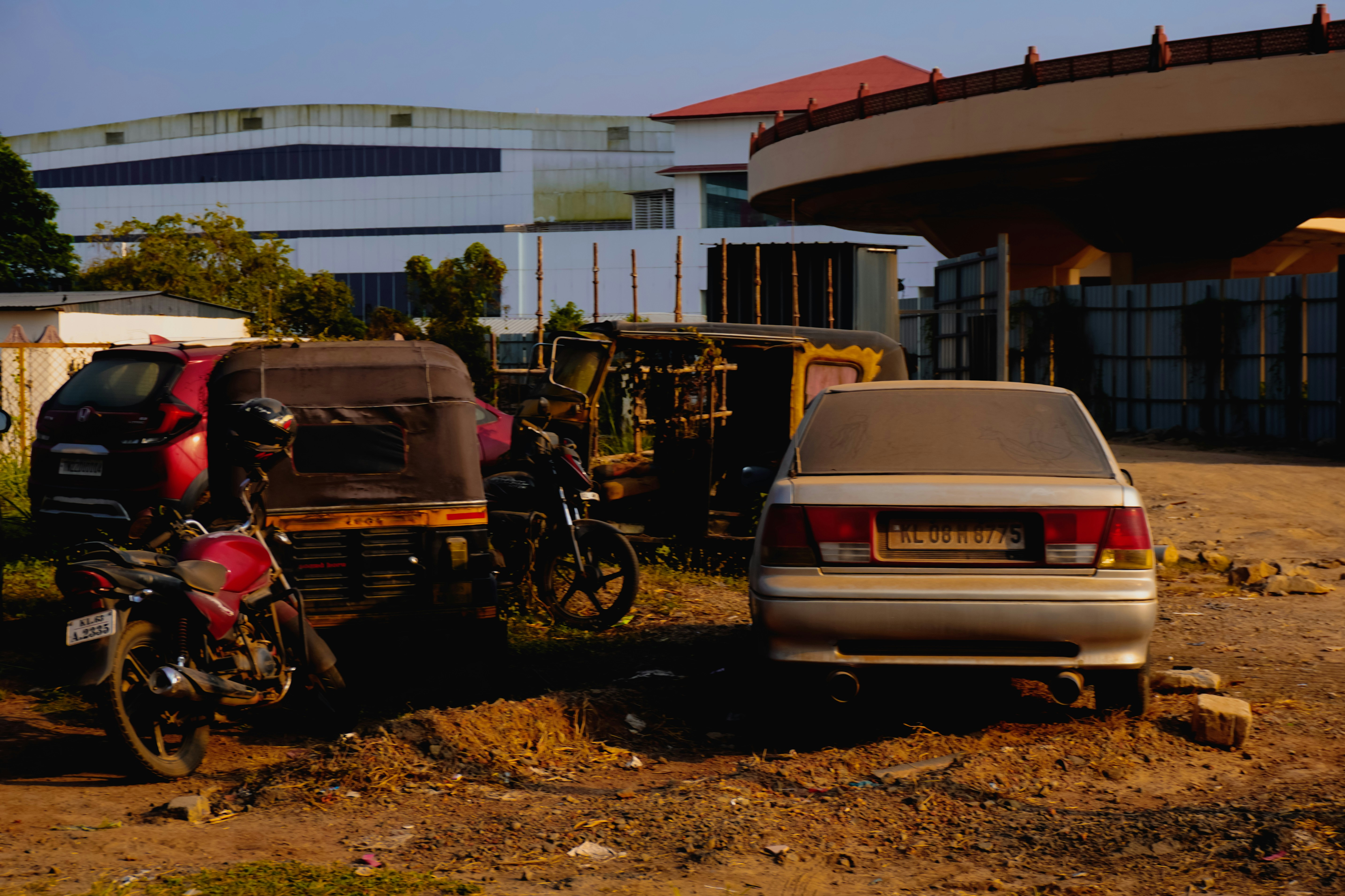 Dusty Car Yard at Twilight: A Silver Sedan Watches Over Motorbikes and Shipping Containers