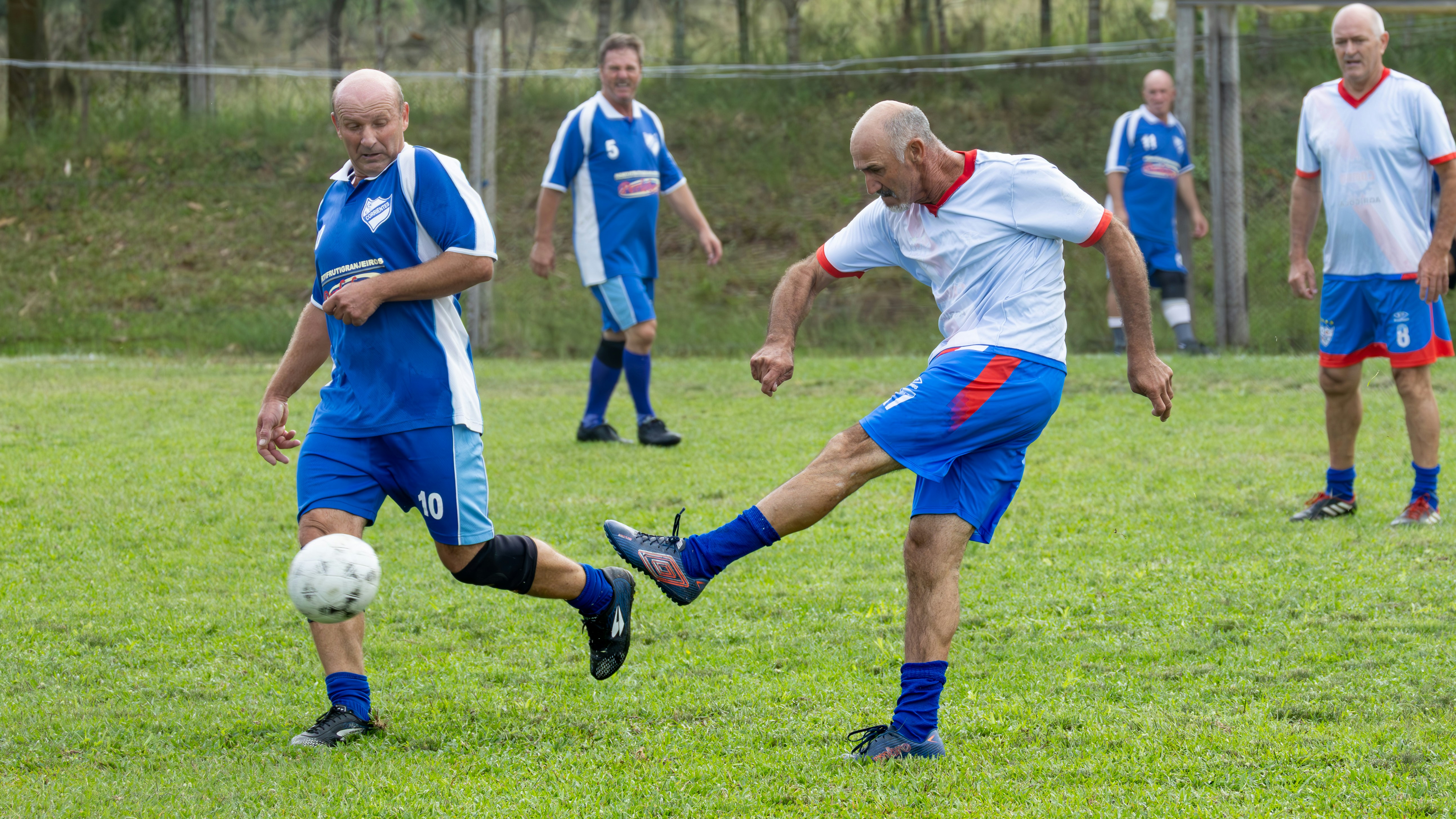Soccer players in blue and white uniforms compete on a grassy field; a player in white and blue is about to strike the ball while teammates observe. A logo and team kits indicate an amateur match setting.