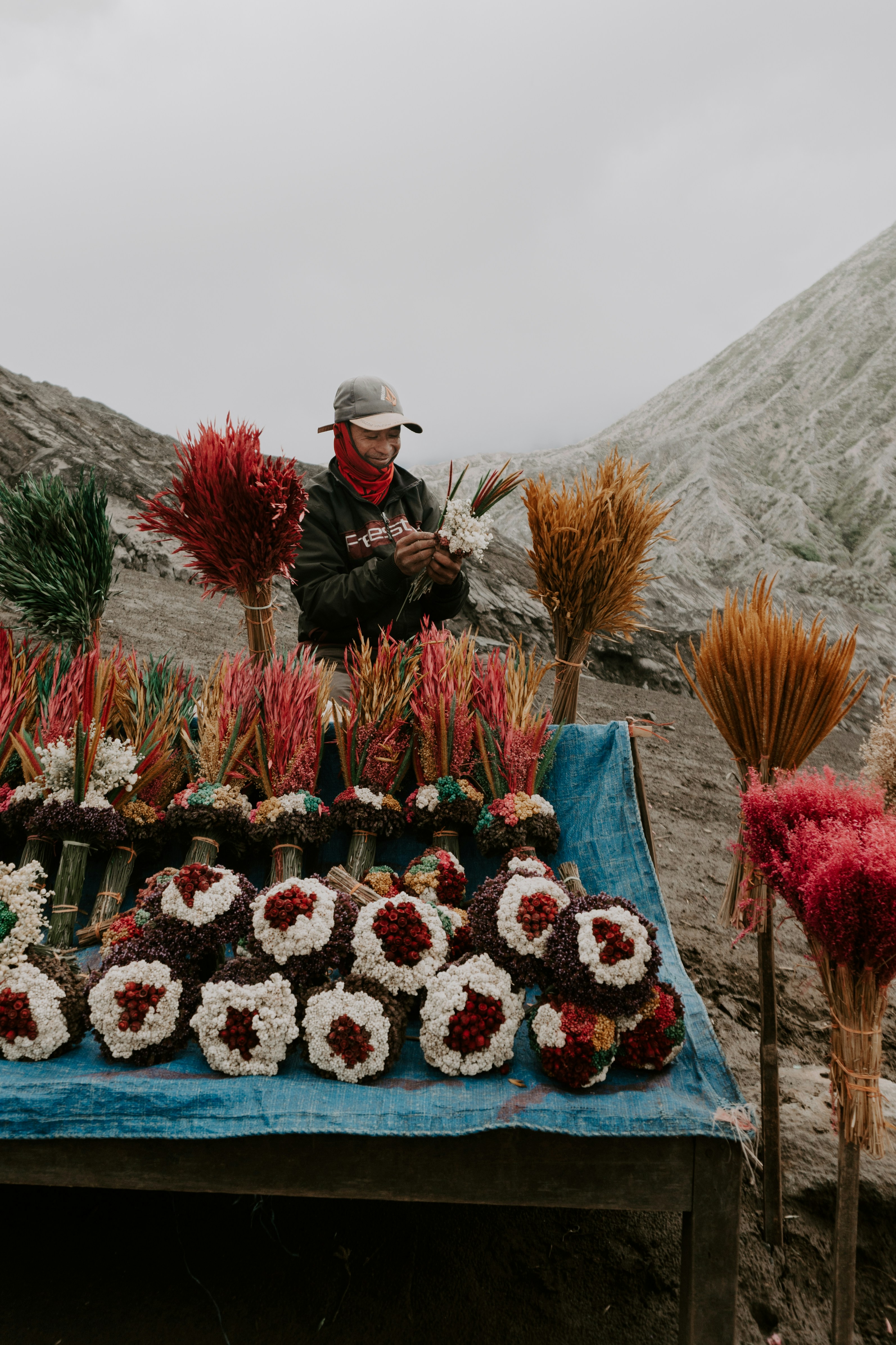 Colorful flower crowns and bouquets arranged on a blue cloth-covered stall with a vendor arranging them, set against barren hillside.