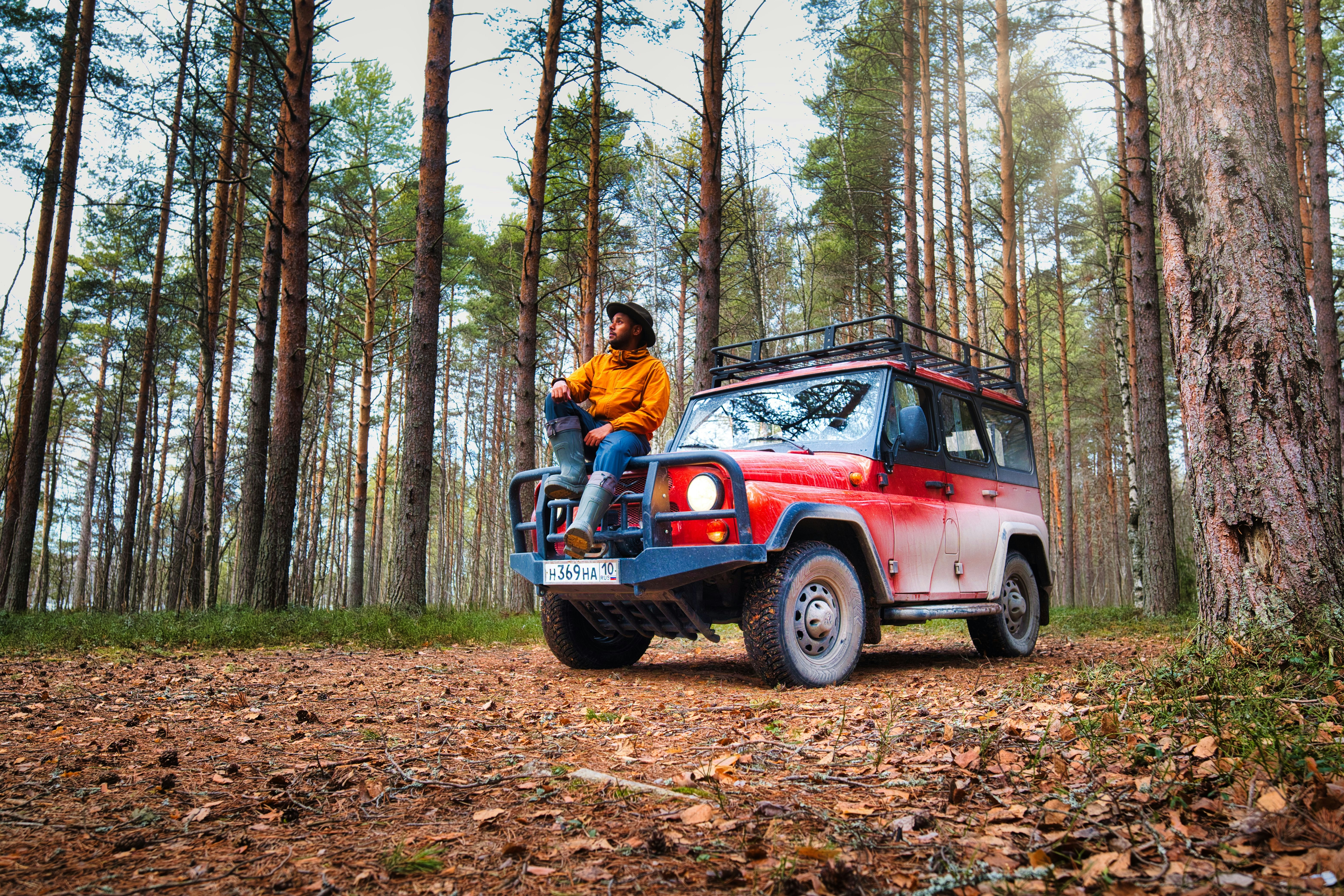 Orange-clad traveler resting on a rugged red SUV in a pine forest