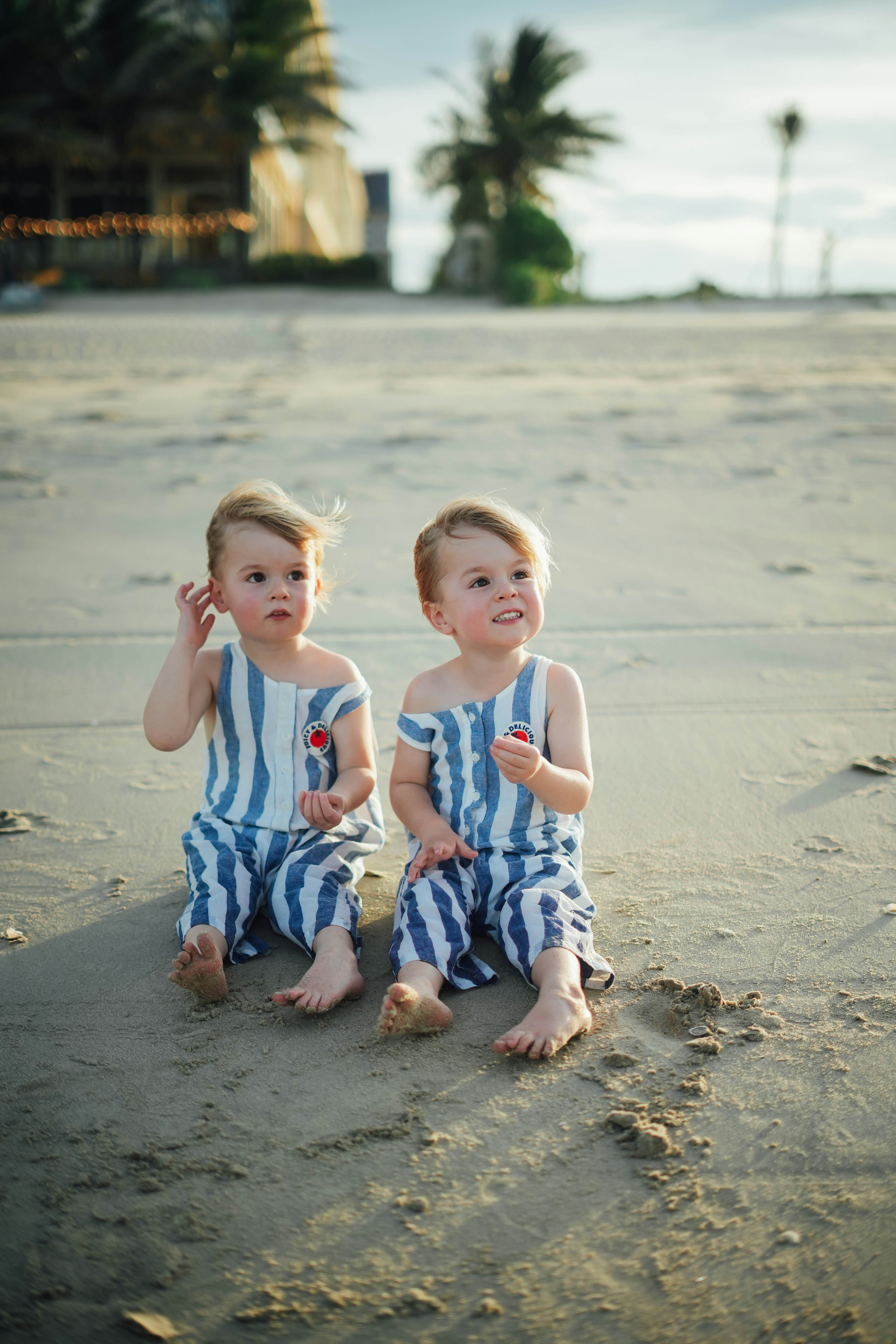 Two Beachward Twins in Striped Overalls Sit in Soft Sand