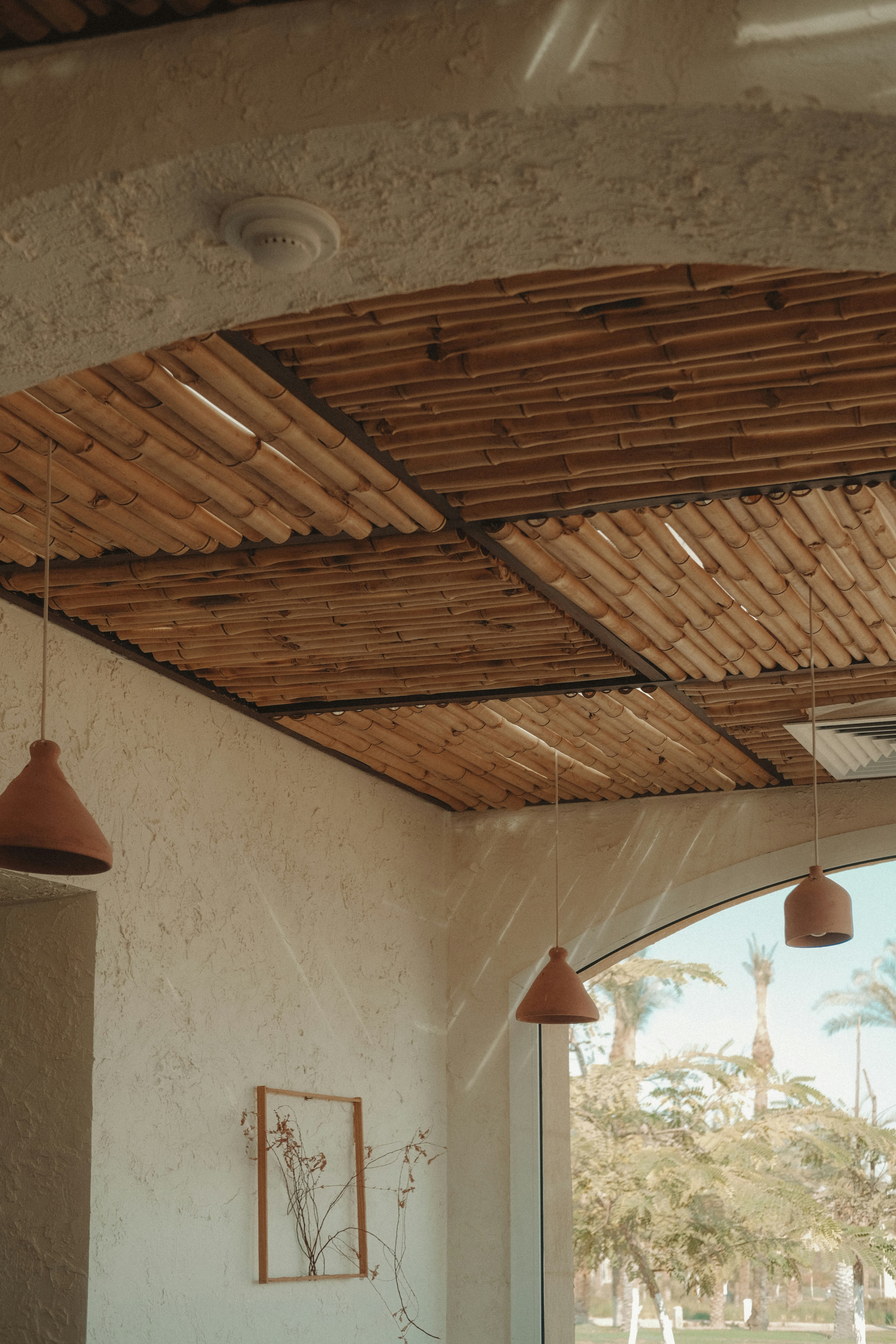 Rustic interior with a bamboo ceiling, hanging pendant lights, and a textured wall; soft daylight filters through a large window, revealing a glimpse of outdoor palm trees.