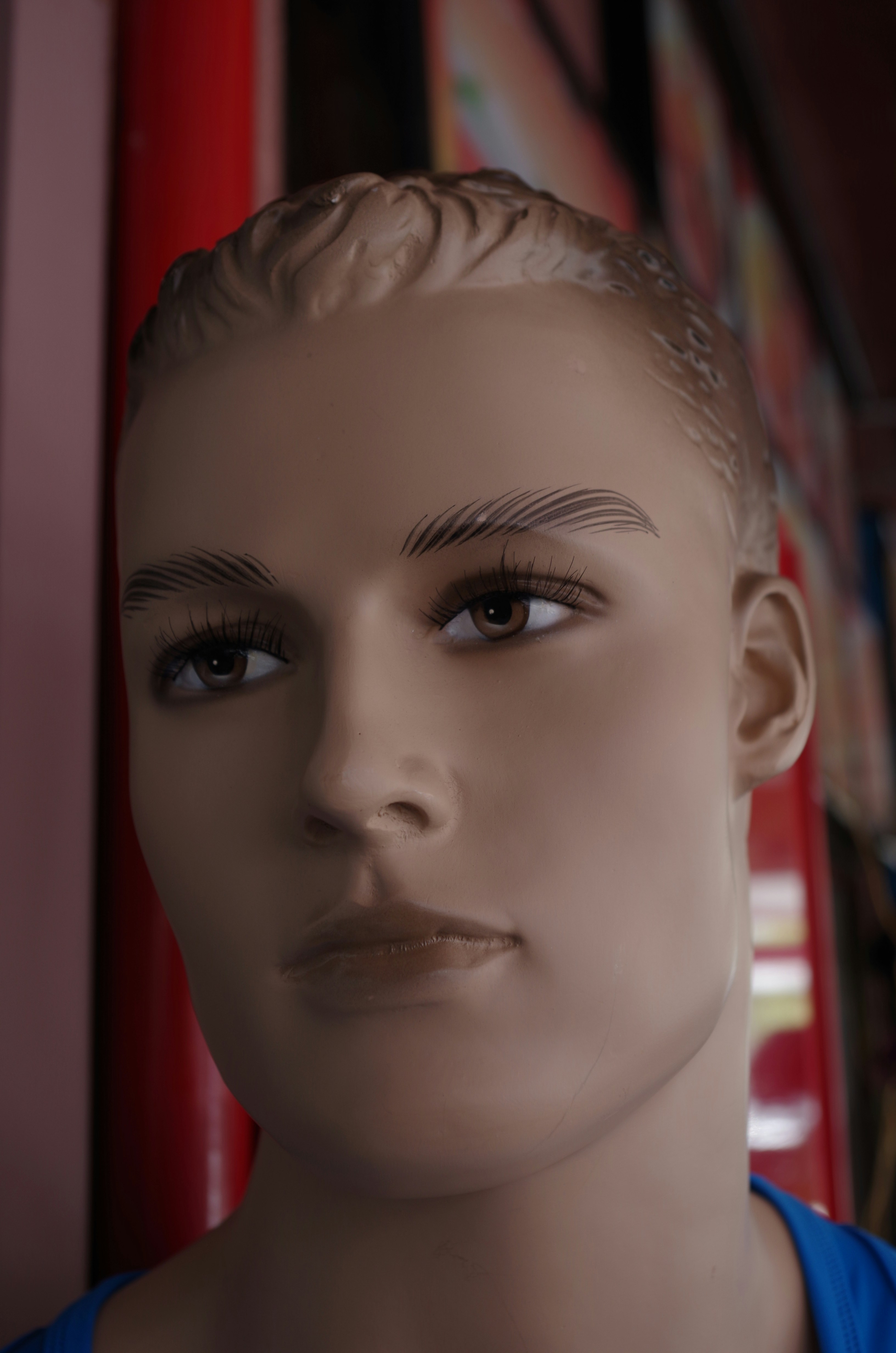 Close-up of a life-size mannequin head with neutral expression against a blurred red and background; a studio style display portrait. Note: this is a mannequin head image.