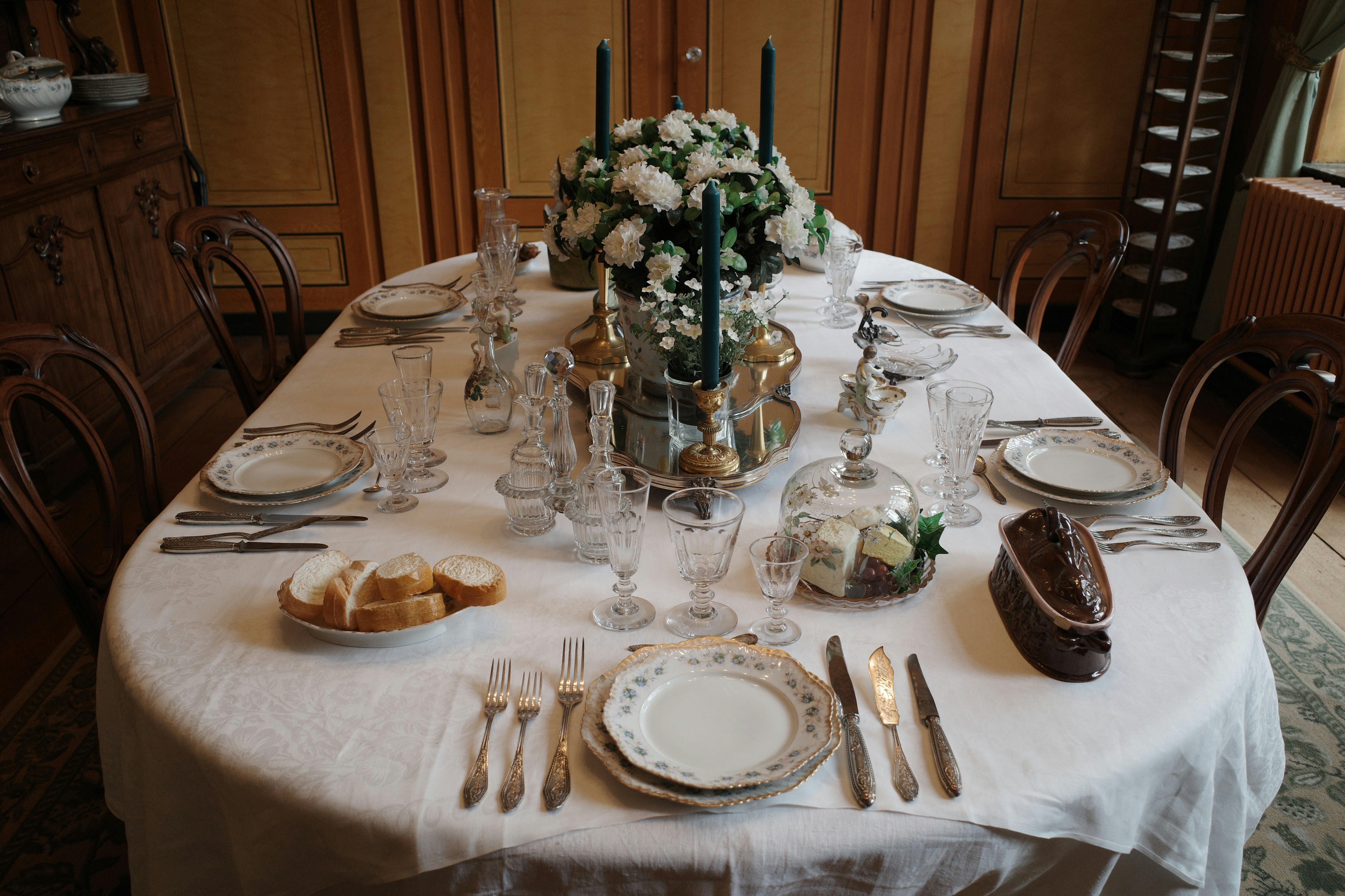 An intricately set dining table featuring a lush floral centerpiece and tall taper candles, with bread, cutlery, glassware, and a bread loaf arrangement arranged for a formal meal. The scene appears as a still-life setup in a refined dining room.
