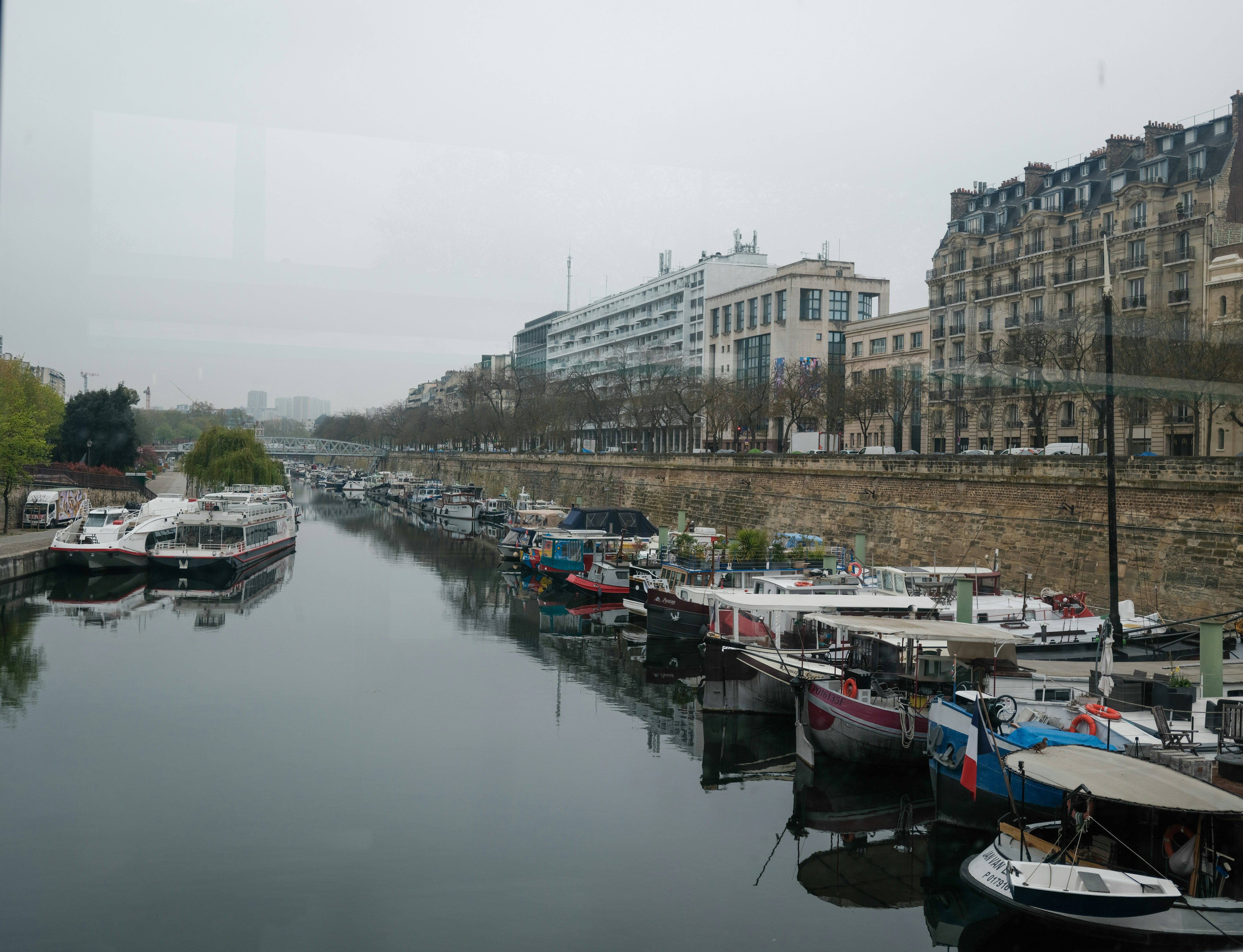 Boats docked along a calm urban canal with historic stone walls and row houses lining the riverbank, captured in soft daylight.