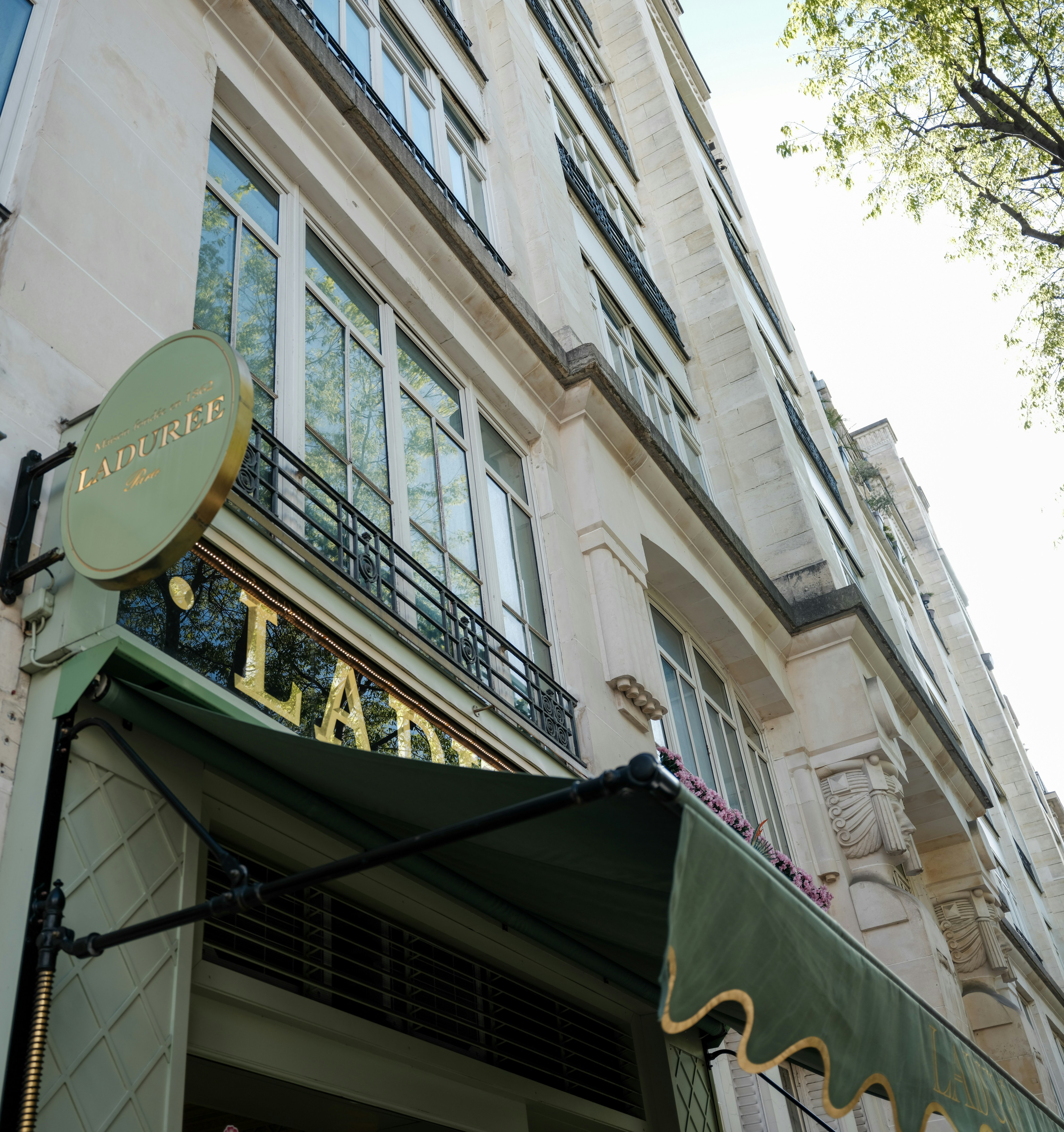 Slightly upward view of a Parisian storefront with a green striped awning and gold signage along an elegant stone building.