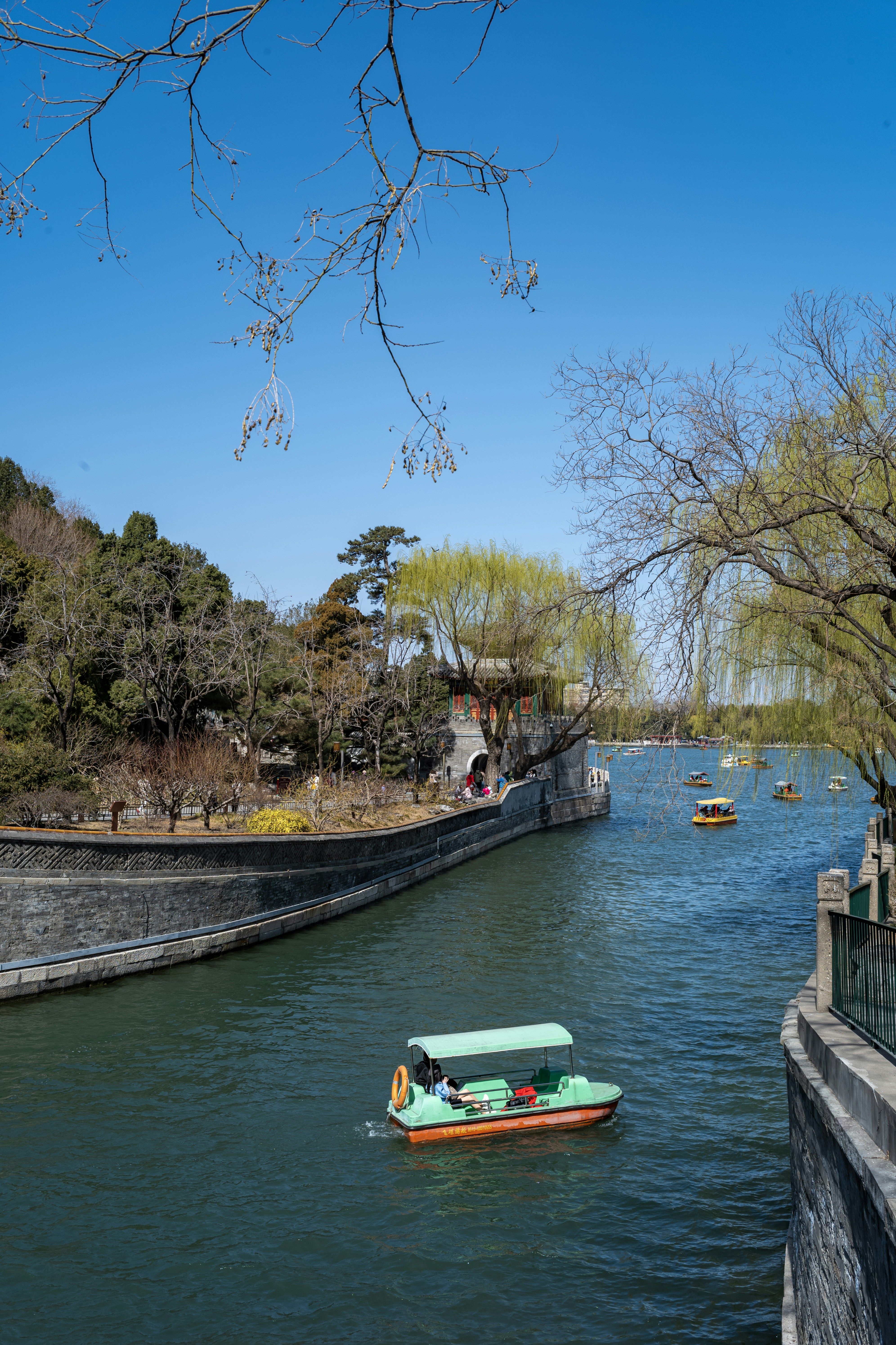 A calm canal lined with stone walls and trees extends into the distance, with a small green boat in the foreground. The scene captures a tranquil day by the water with a clear blue sky and light foliage.