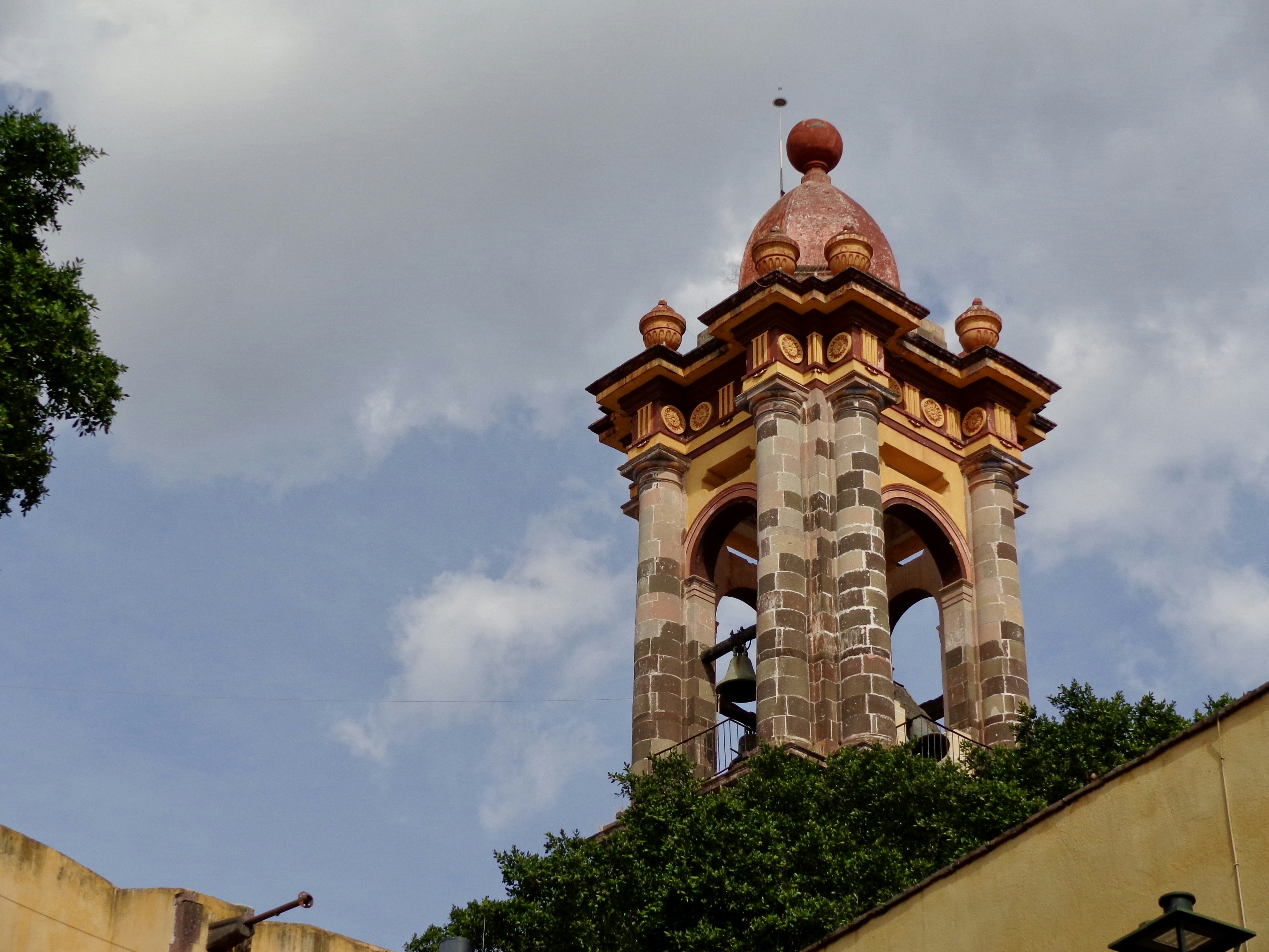 Stone bell tower with decorative brickwork rises above green foliage against a partly cloudy sky.