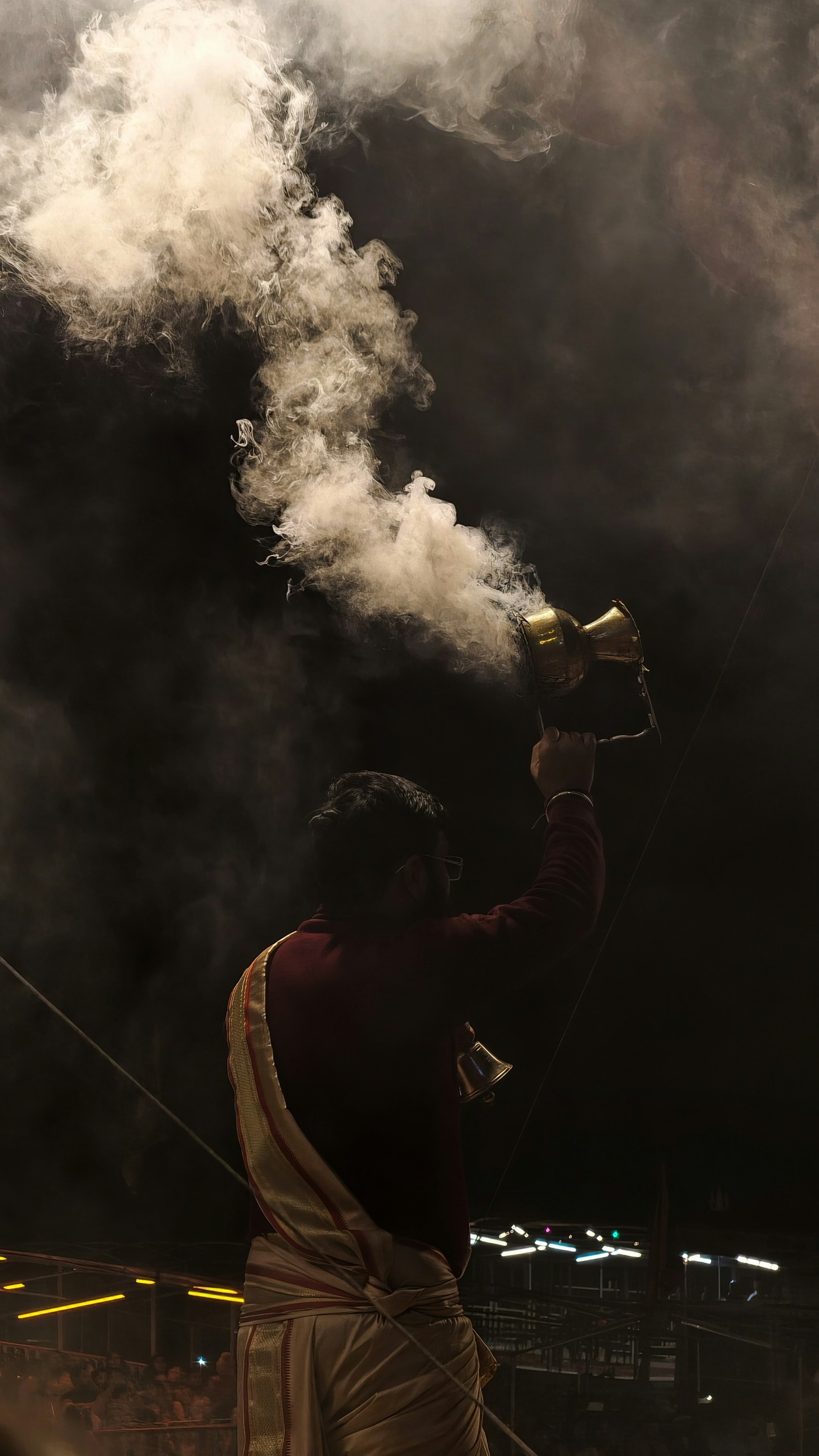 A silhouetted man in traditional attire raises a brass horn into a cloud of smoke against a dark backdrop, with stadium lights faintly glowing in the distance.