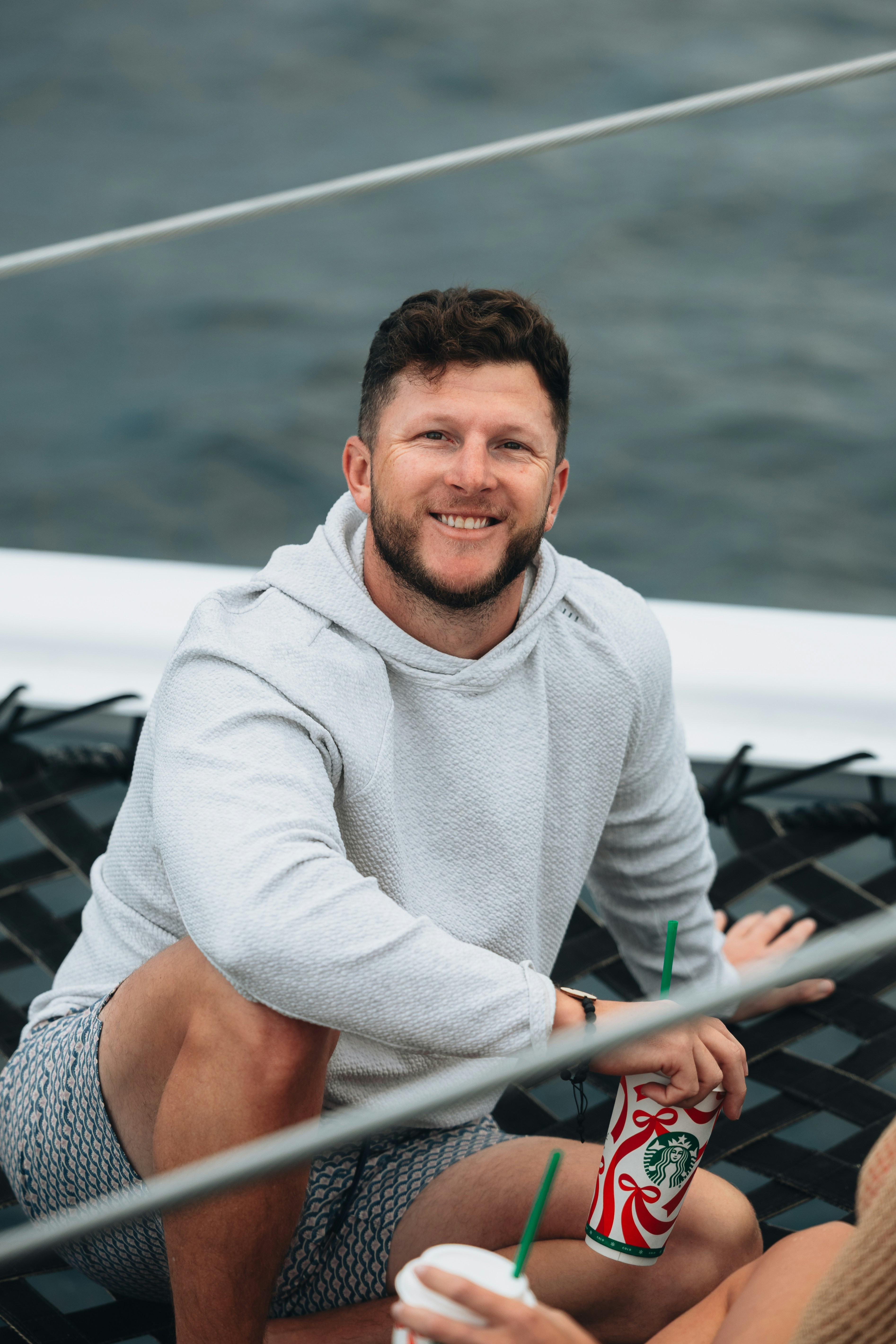 Smiling man in light gray hoodie on a boat, holding a Starbucks cup